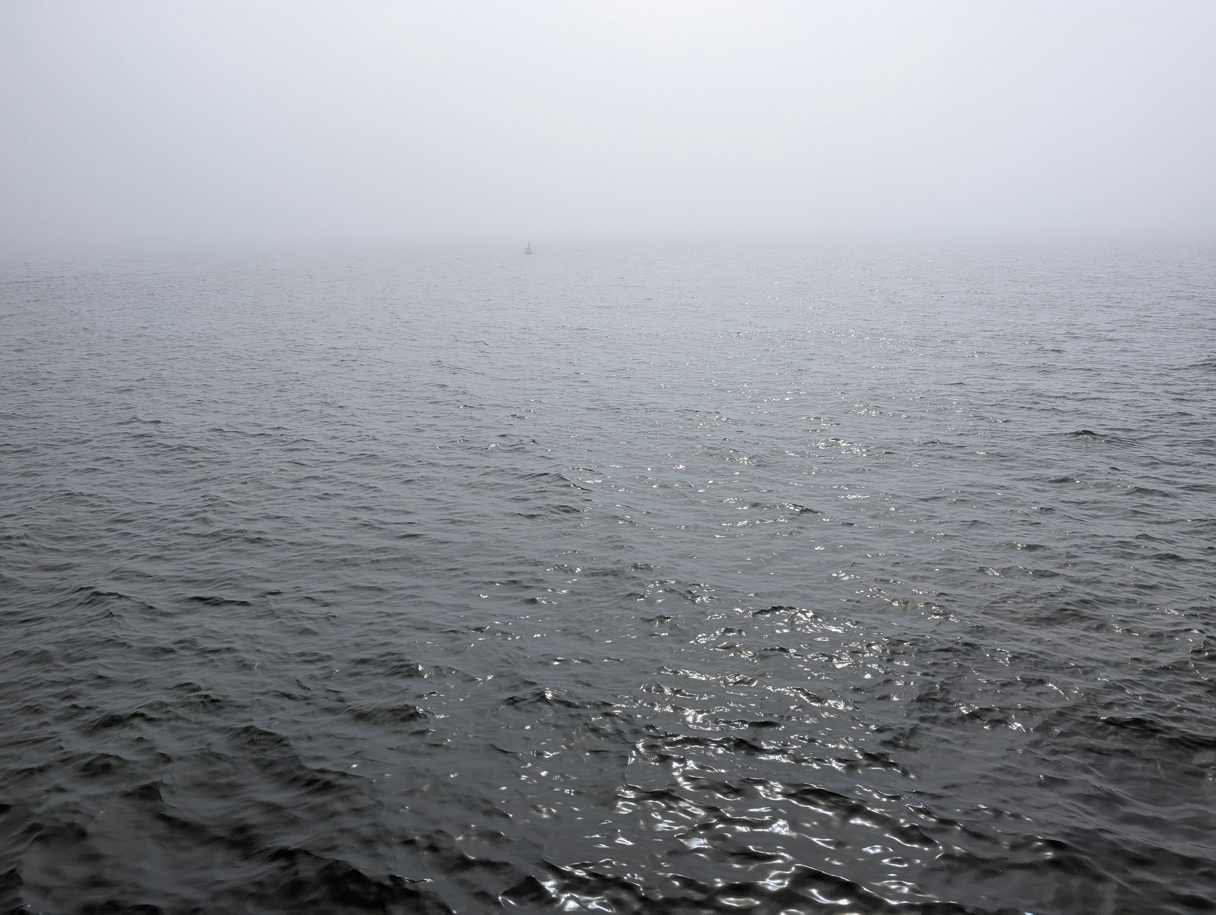 Muted seascape with a faint horizon and glimmering sunlight on gentle waves; the image emphasizes vast openness and atmospheric haze, with no distinct foreground subjects.