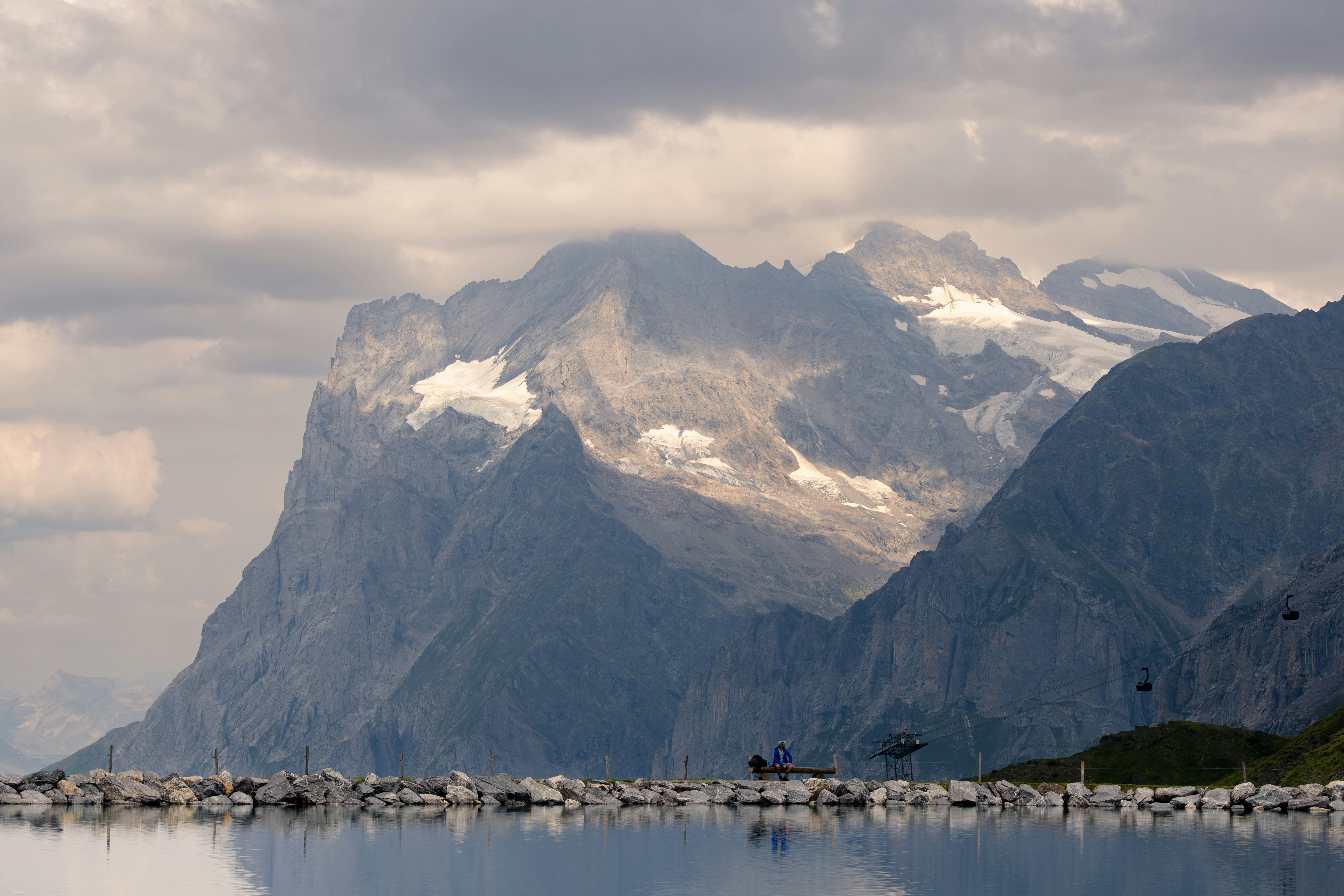 Snow-dusted alpine peaks rise above a calm lake, with a thin rocky breakwater and a hint of human presence grounding the vast landscape.