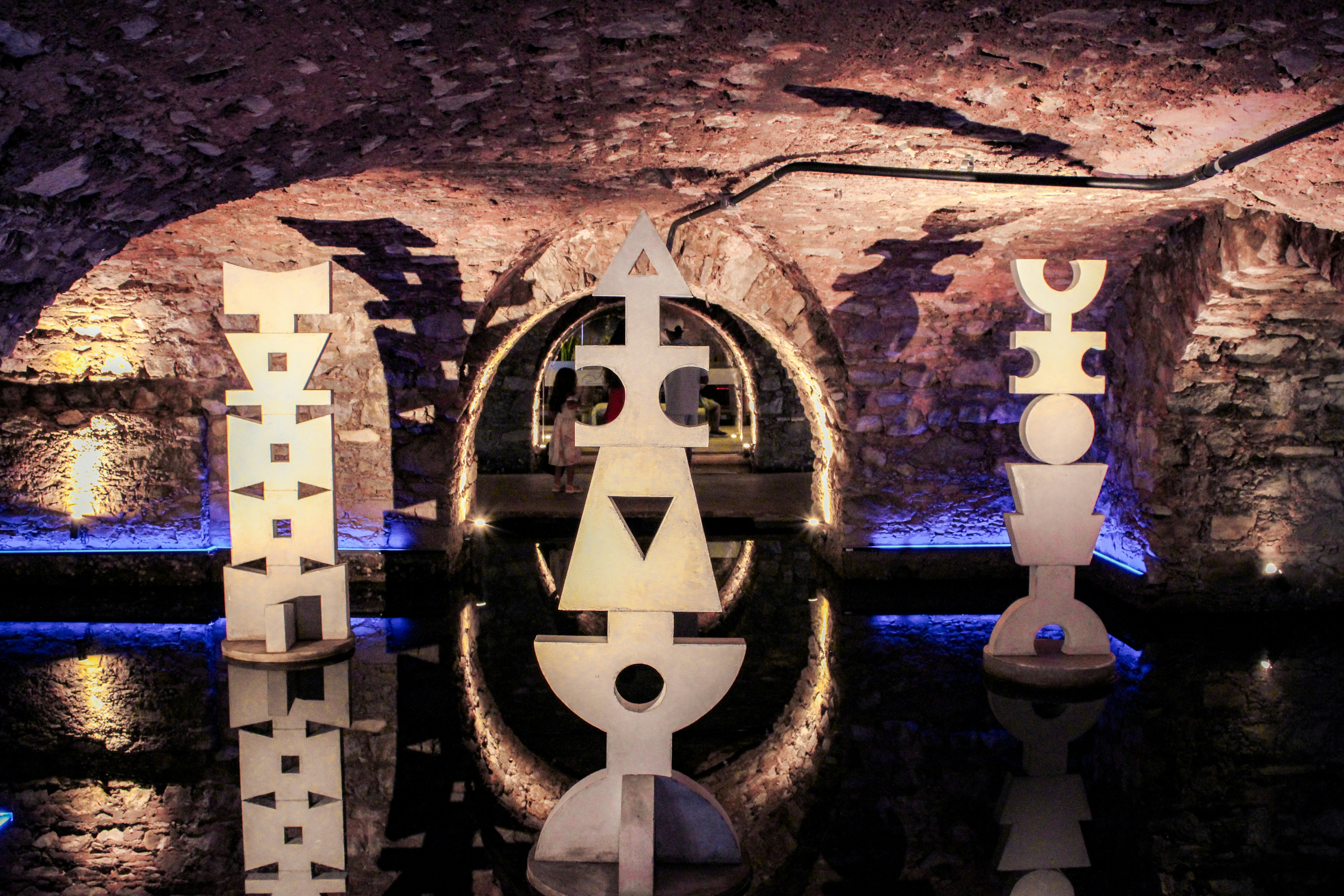 Symmetrical geometric sculptures rise from a reflective pool beneath an ancient stone arch, illuminated by warm and cool tones. Abstract forms and their mirrored silhouettes create a tranquil, architectural tableau.