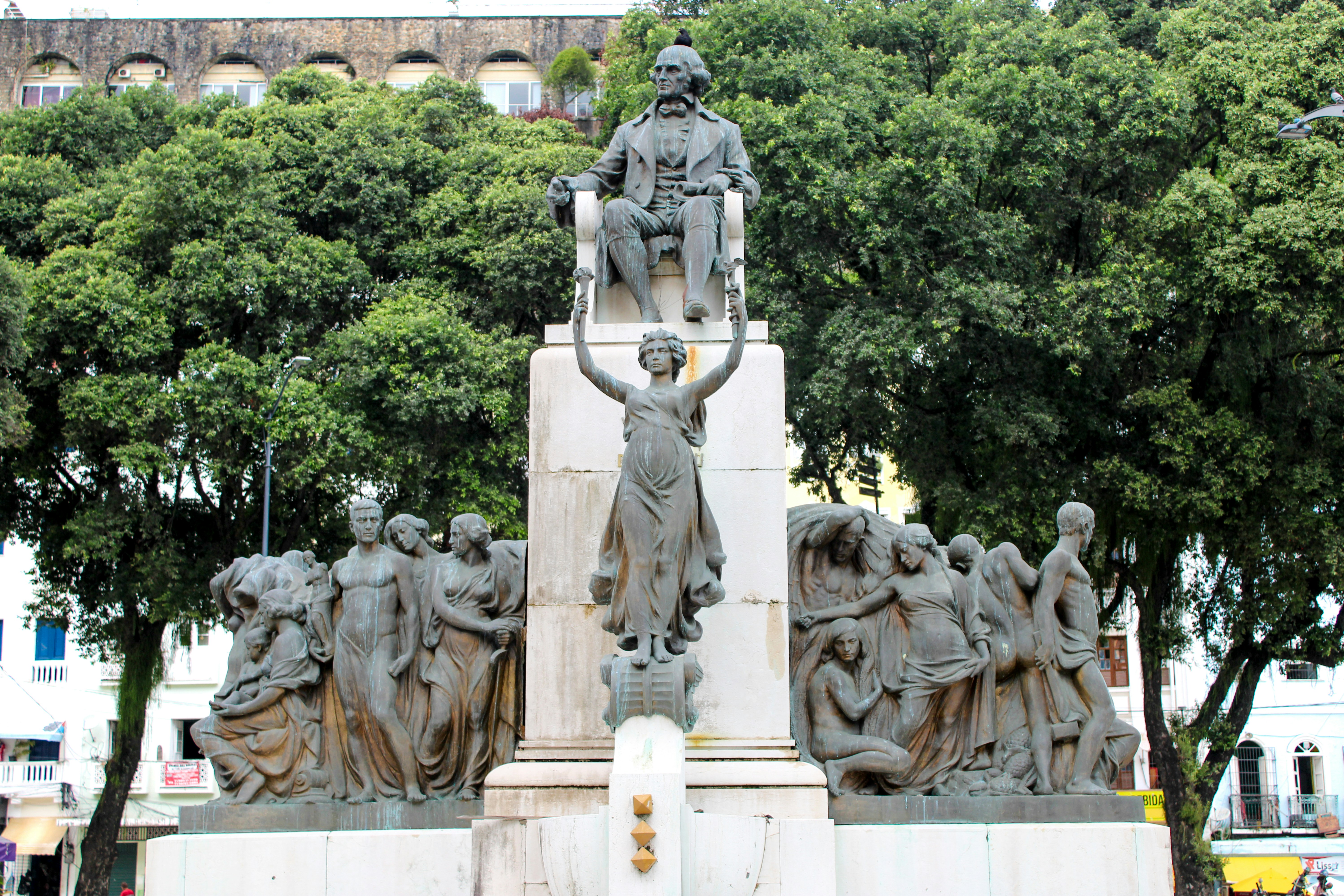 Statue Ensemble: Central Pedestal Framed by Sculpted Figures in a Lush Urban Square
