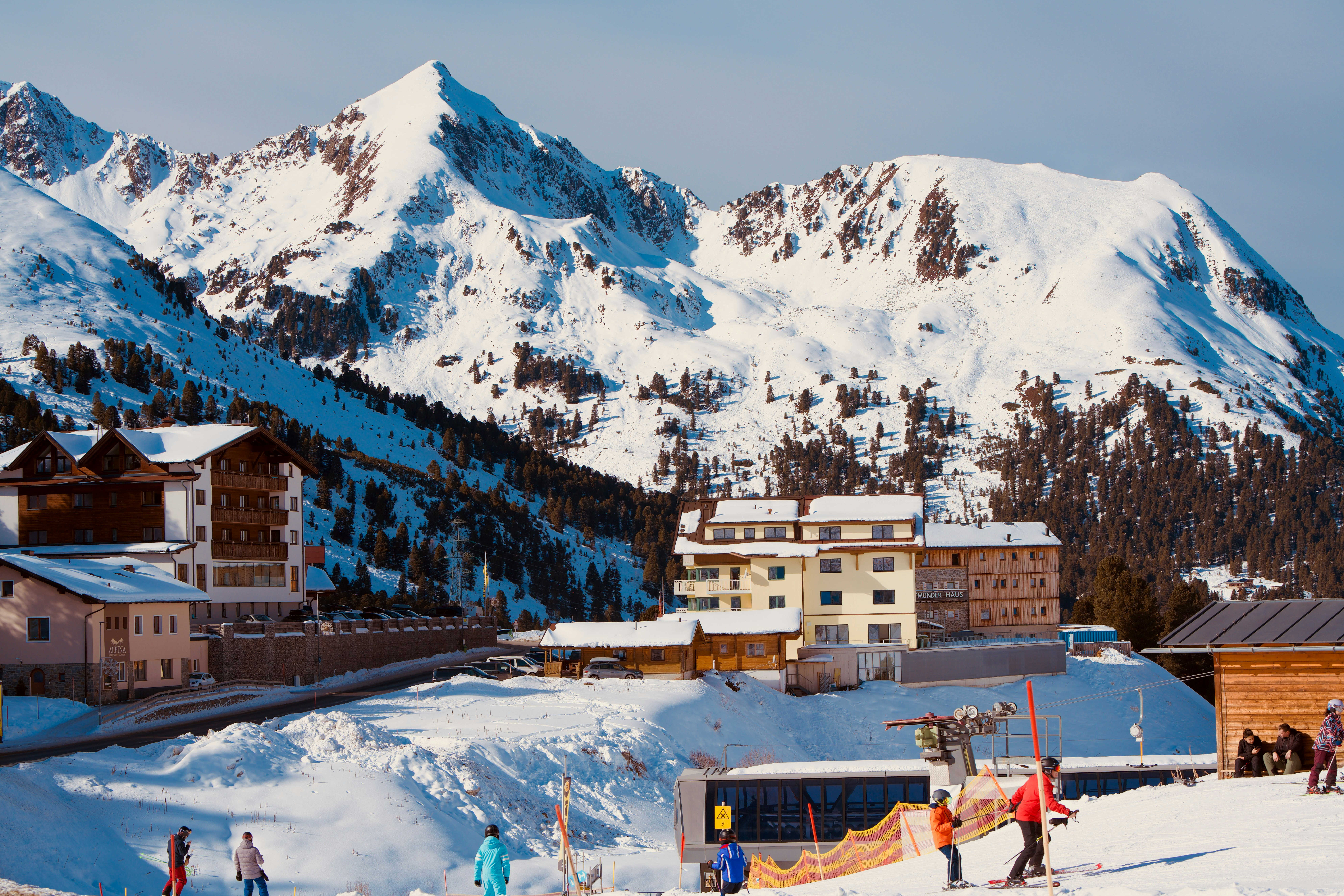 Snow-covered alpine village with pastel buildings at the base of rugged mountains; skiers and a small slope in the foreground.