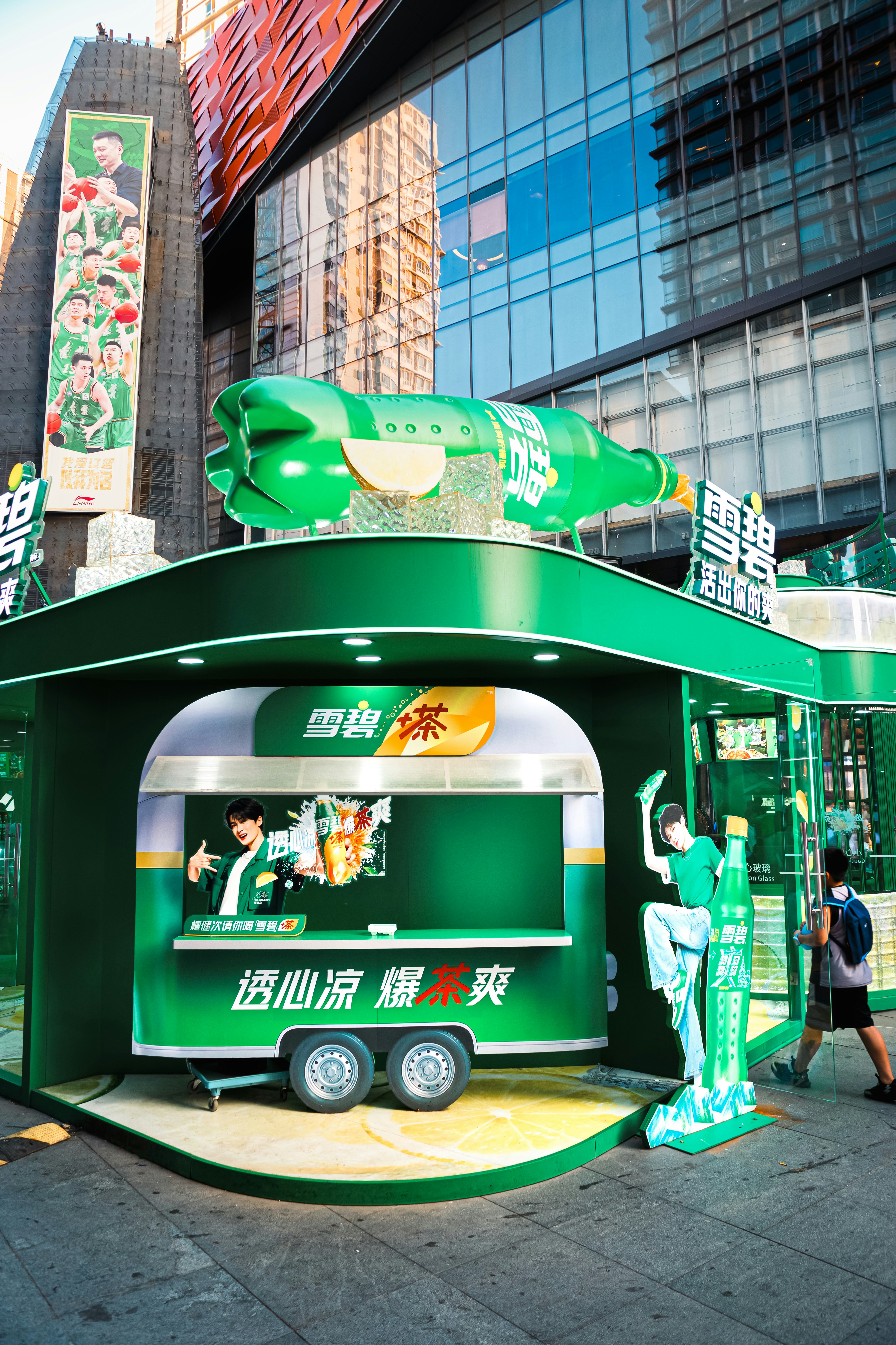 Green beverage promotional stand with a large inflatable bottle atop a curved kiosk in an urban plaza. A life-sized mascot and a video display anchor the bright green setup beside glass-fronted buildings.