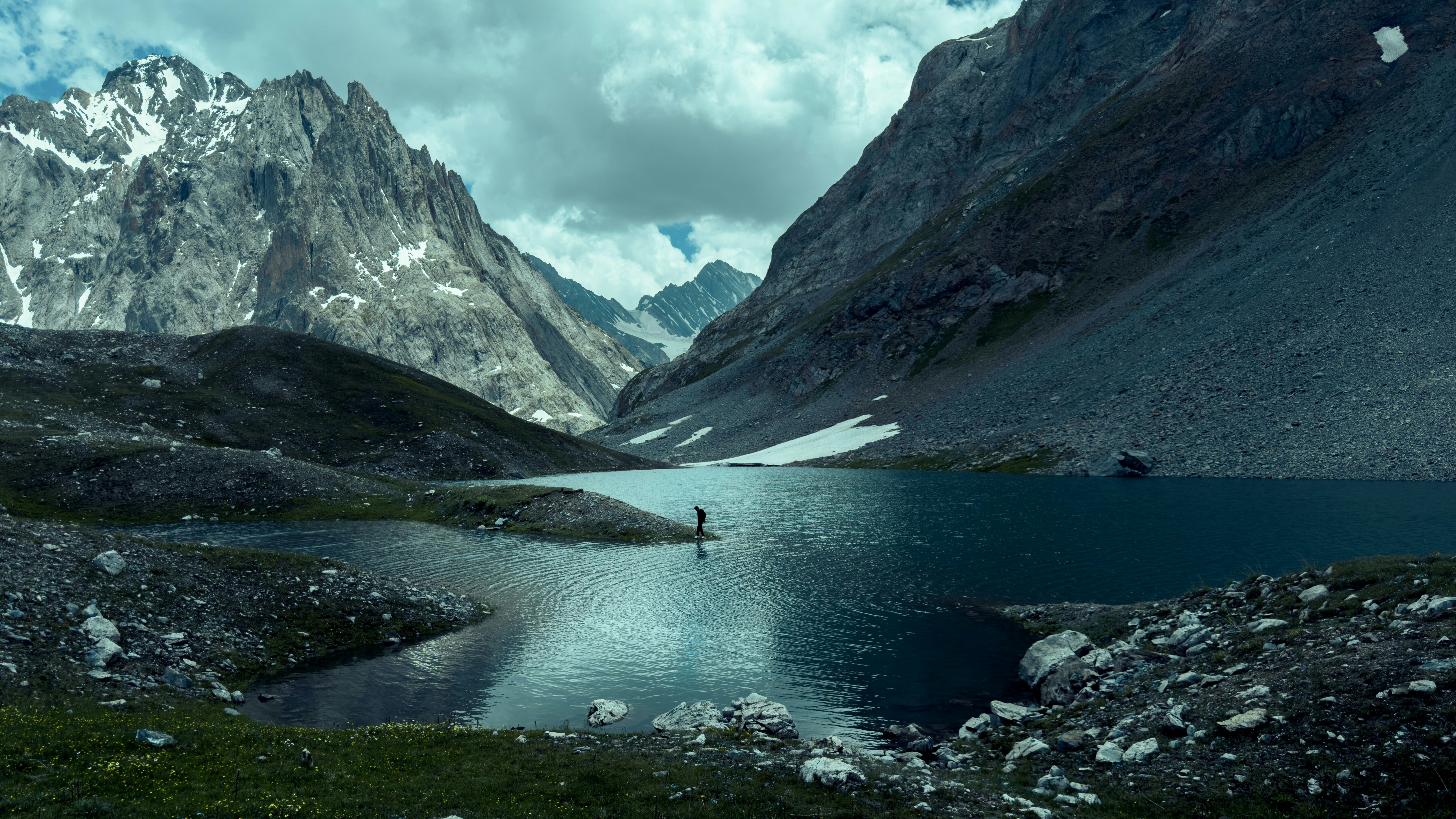 Crescent alpine lake reflects rugged peaks and a snowy slope, with a lone figure emphasizing scale; a natural landscape with stark rock, water, and distant ice. (Photo is a landscape scene.)