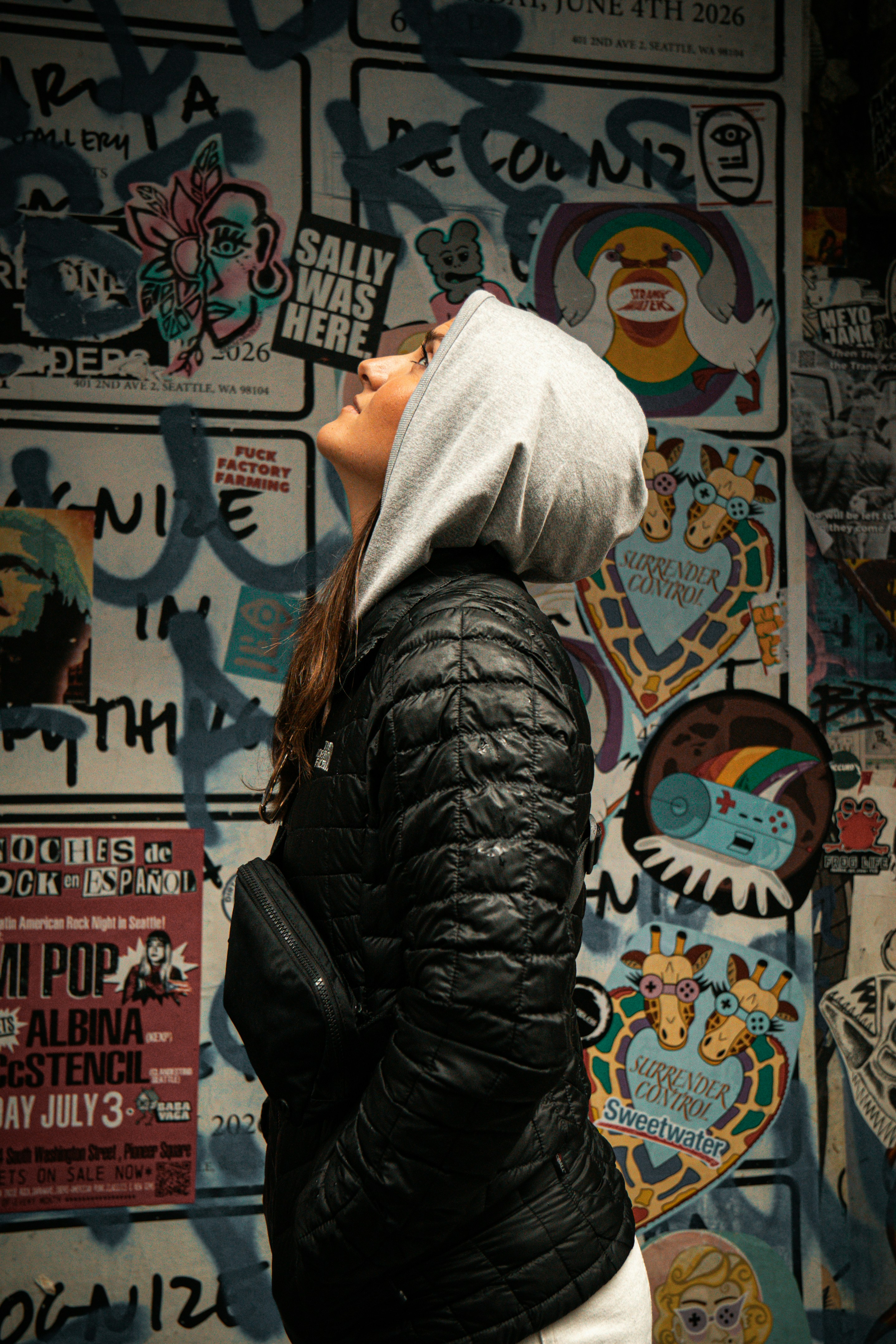 Upturned gaze under a gray hood amid a wall of urban art