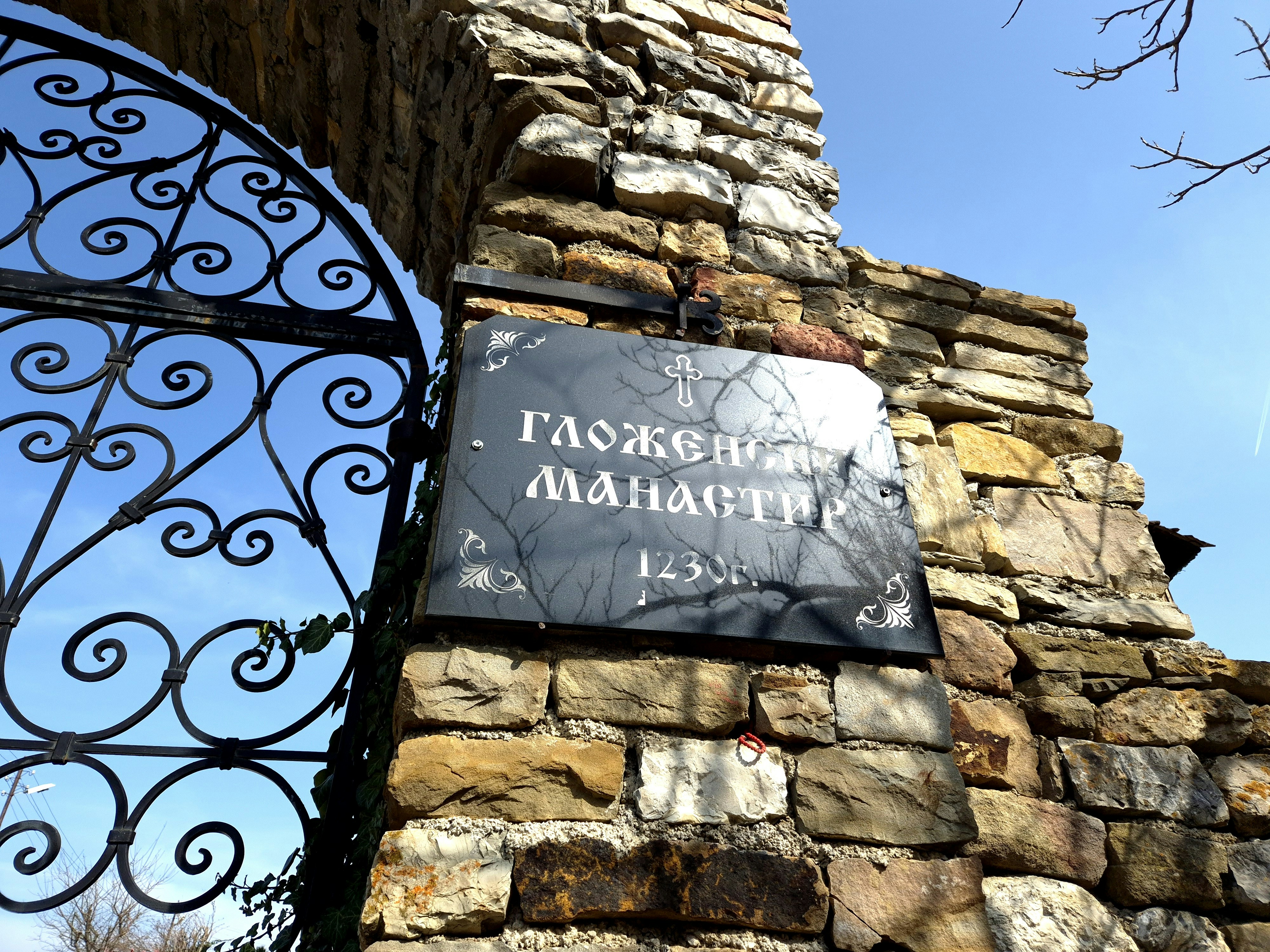 Stone entrance wall of Glozhene Monastery with its engraved sign dating the foundation to 1230, framed by a wrought iron gate. The medieval Orthodox monastery of Saint George sits in the Teteven area of the Balkan Mountains.