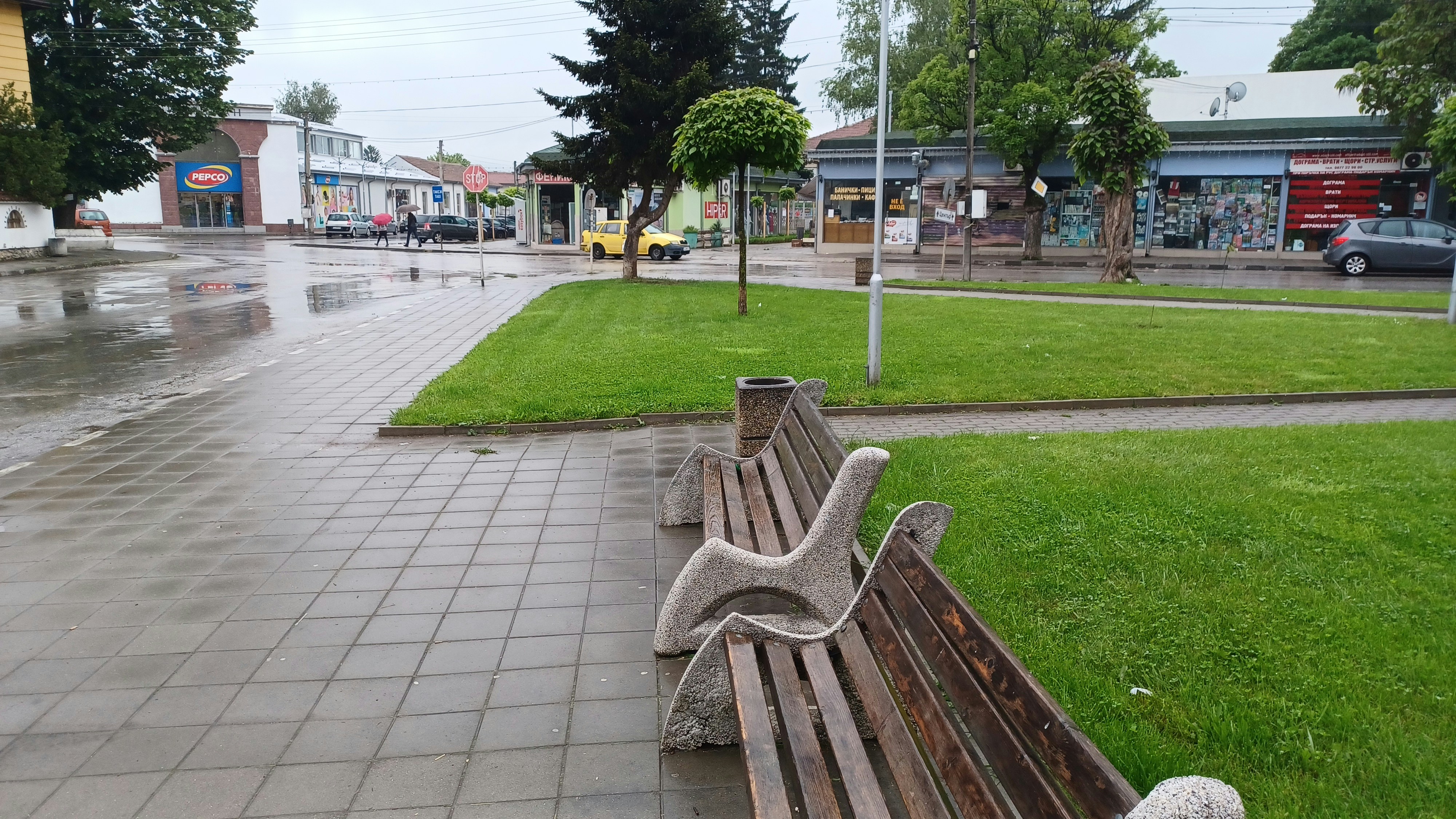 Overcast town square: rain-washed benches and a quiet, green edge