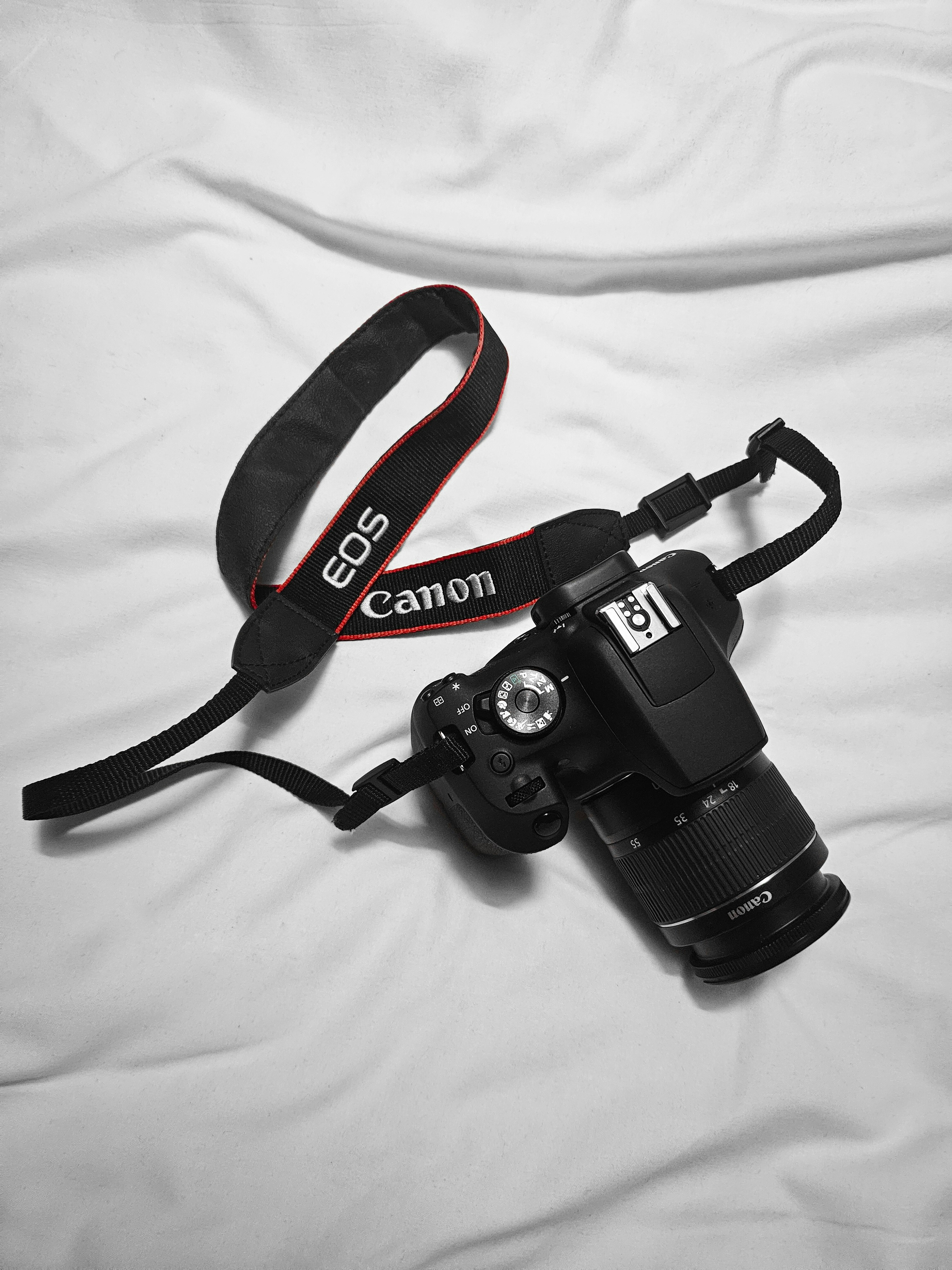 Canon camera resting on soft white fabric, strap draped nearby, monochrome tone emphasizing details of the lens and controls.