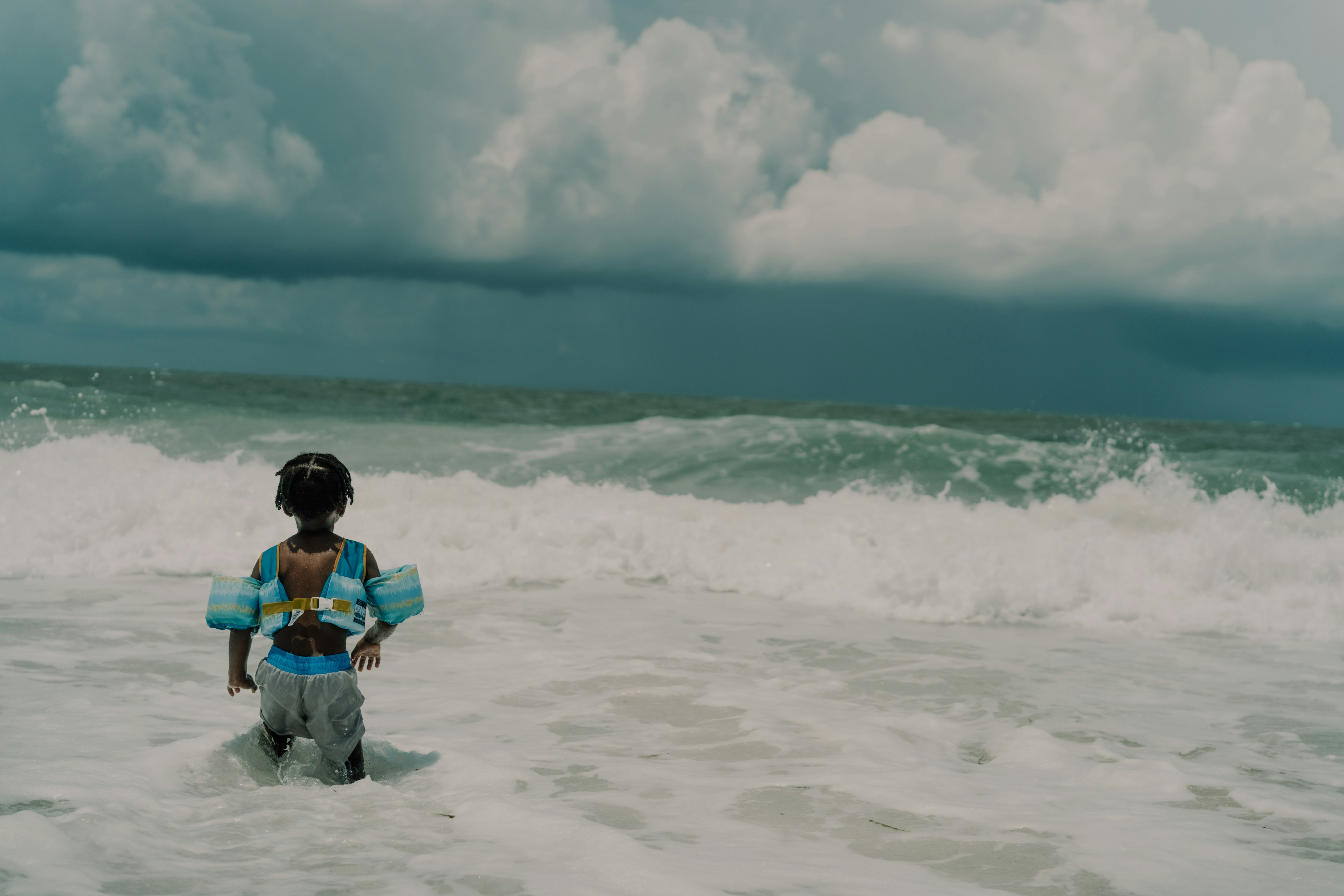Child in a turquoise life vest stands at the edge of rolling surf