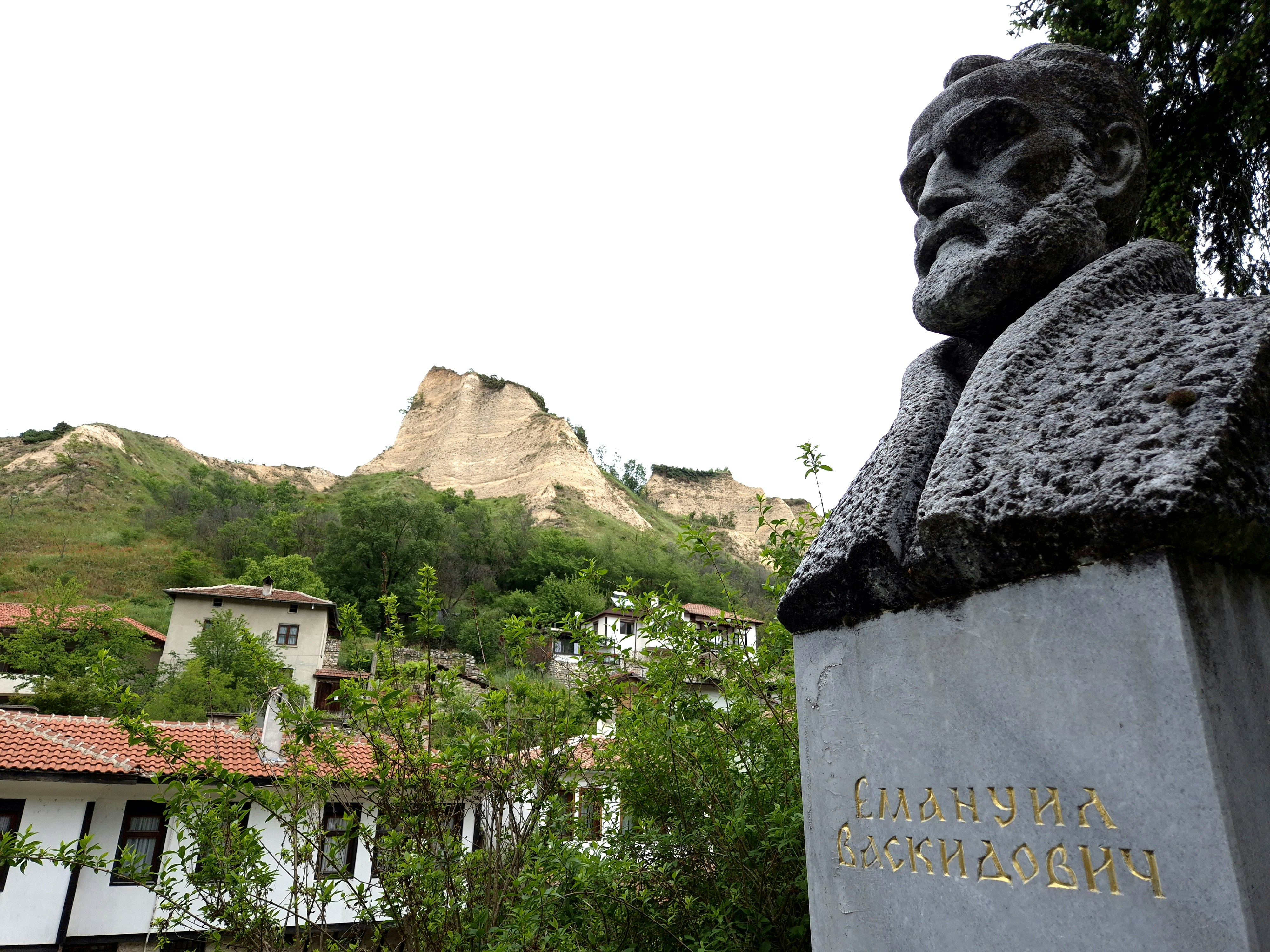 Statue head in the foreground with a carved inscription, overlooking a village and eroded rock formations in the background.