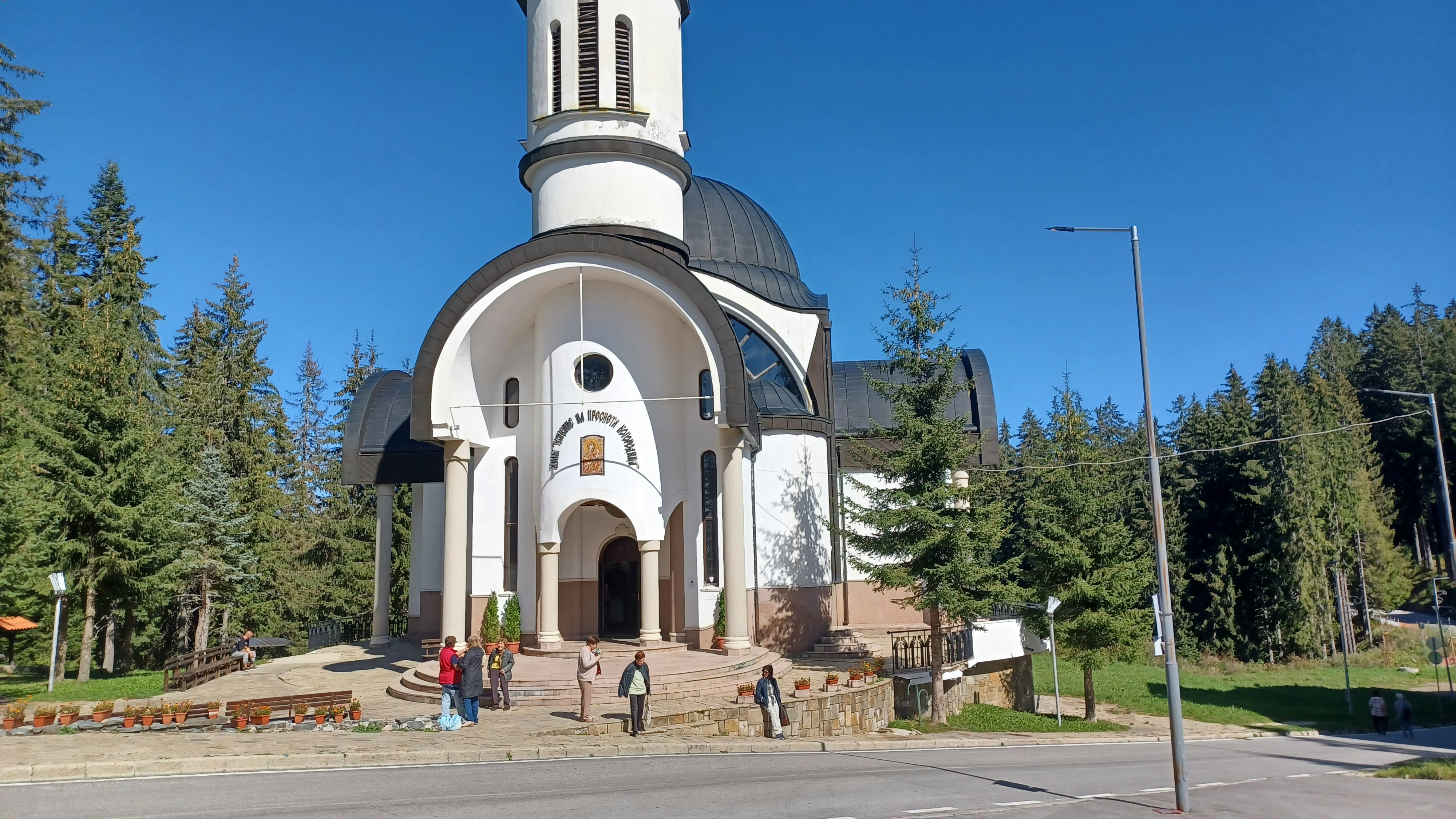 A small white chapel with a dark dome sits at the edge of a pine forest, visitors near the entrance on a sunny day.