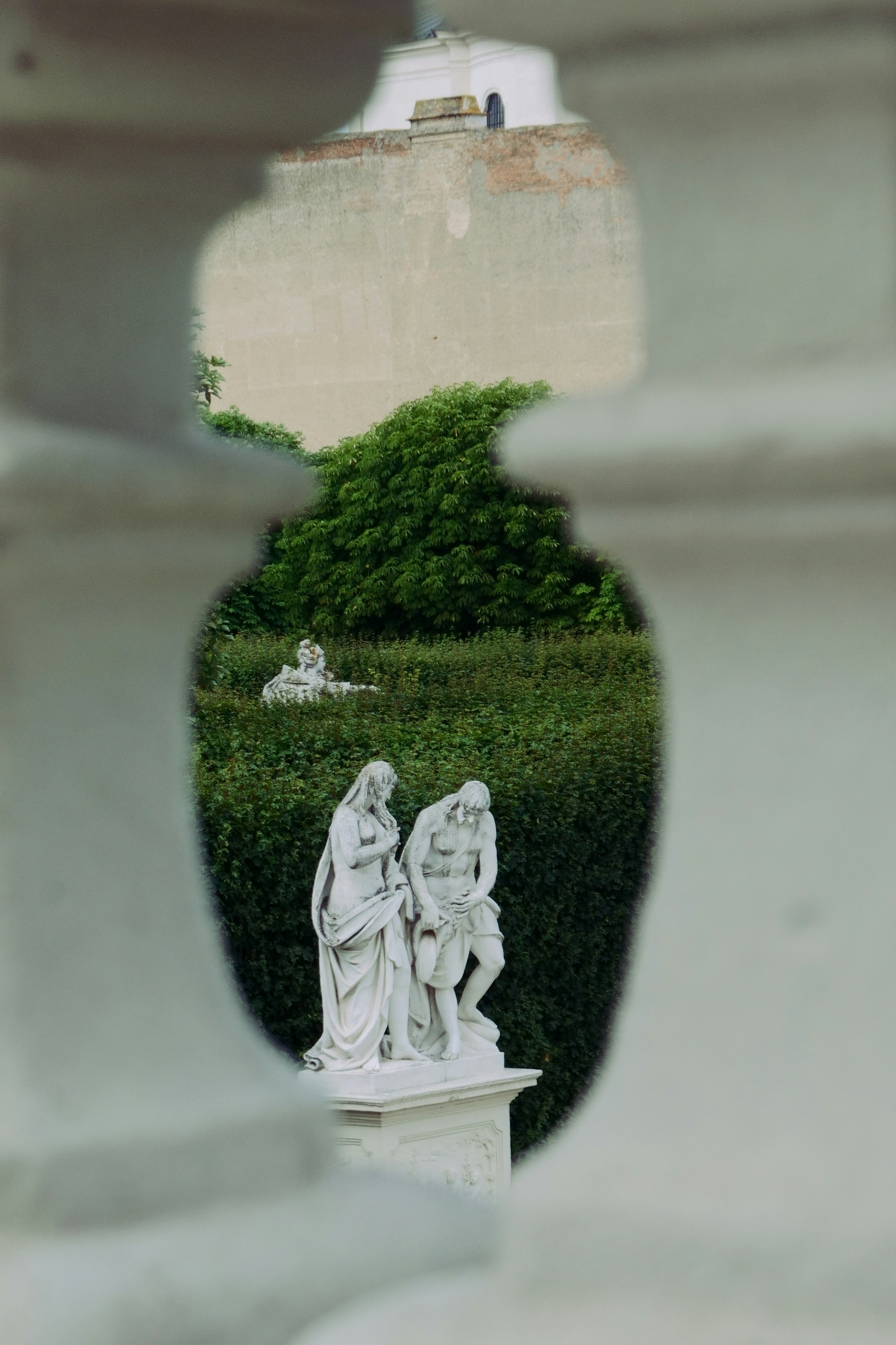 Carved statues sit in a manicured garden framed by foreground balustrades, with greenery and a distant wall in the background.