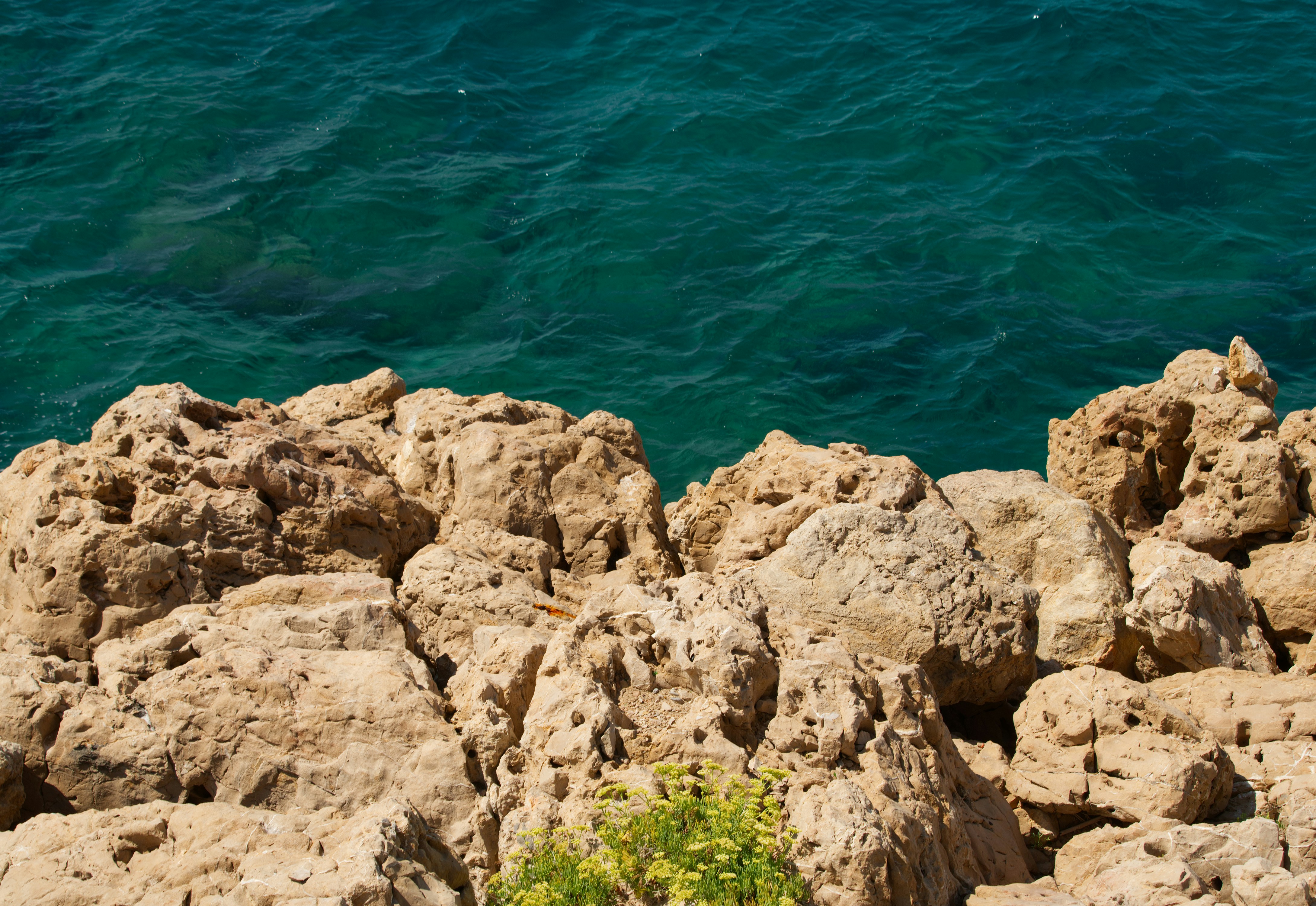 Sunlit beige rocks edge a vivid turquoise sea, captured from above to emphasize texture and contrast against calm water.
