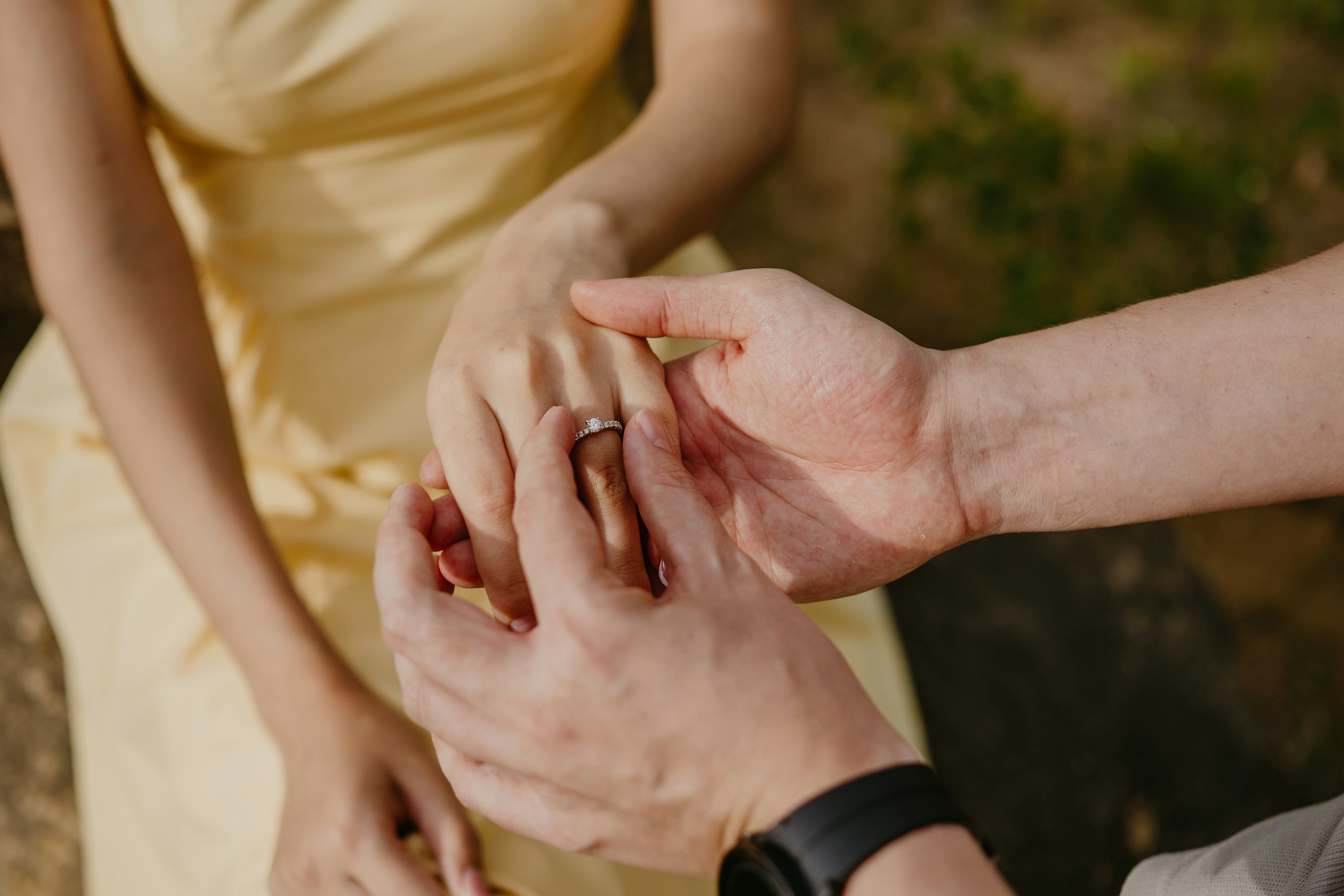 Two hands, one with a ring, intertwine in a warm outdoor setting, capturing a candid moment of commitment amid subdued background. A close-up of the clasping hands emphasizes texture and emotion.