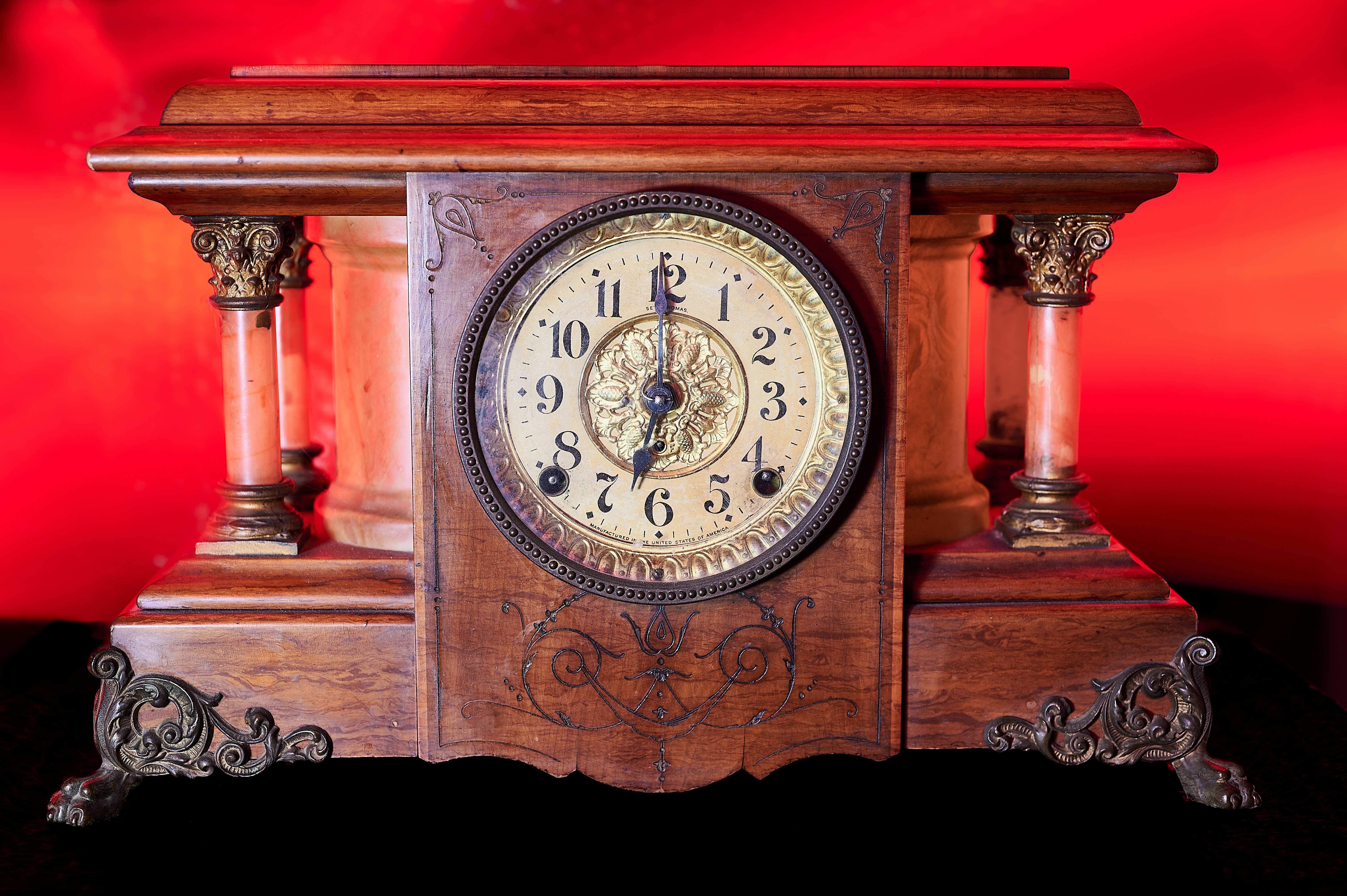 Antique wooden mantel clock centered on a red background, featuring intricate metal embellishments and a decorative dial with ornate hands.