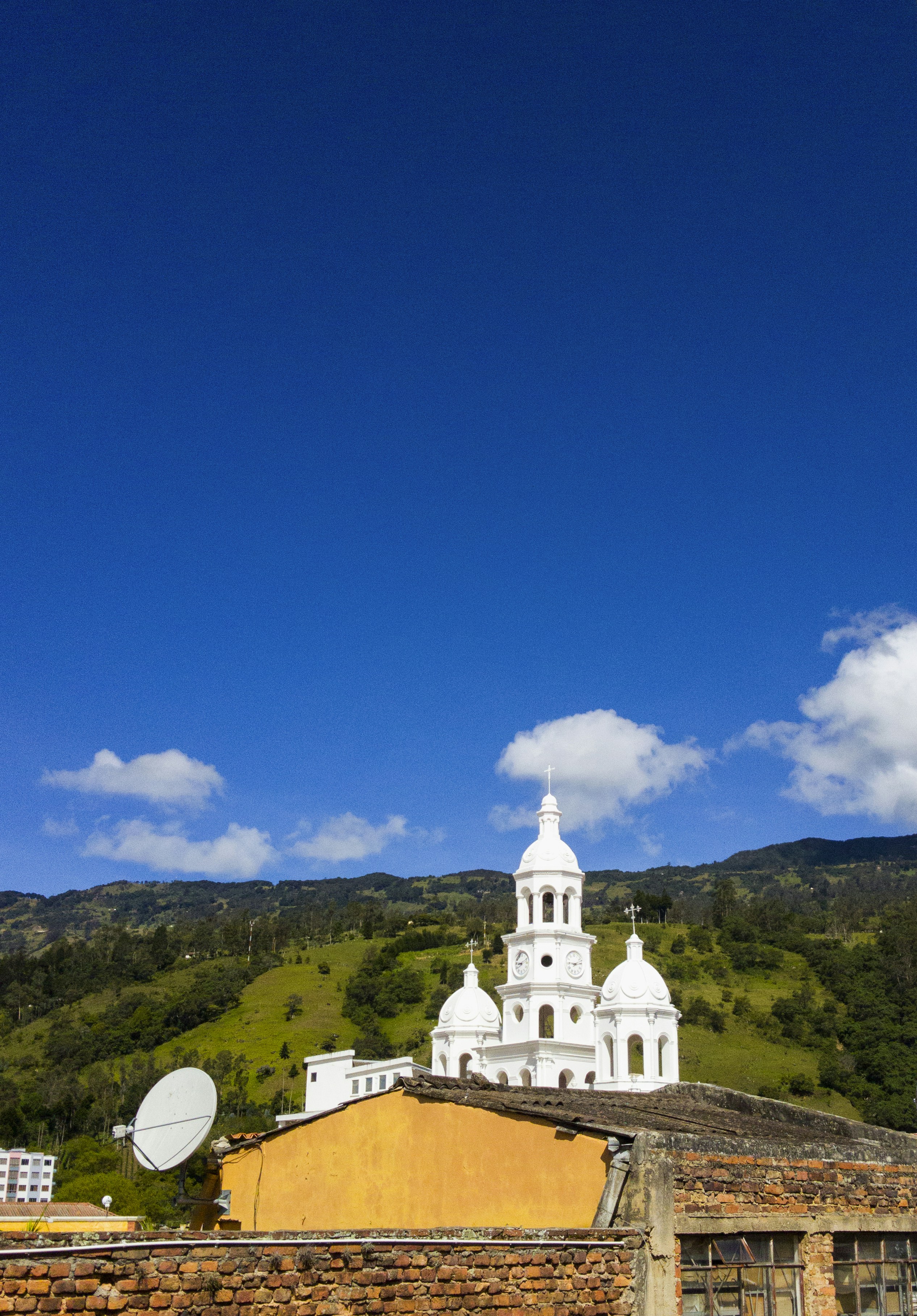 Twin white domed towers rise above a sunlit orange rooftop against a rolling green hillside and clear blue sky. A satellite dish sits at the foreground, adding a hint of urban texture.