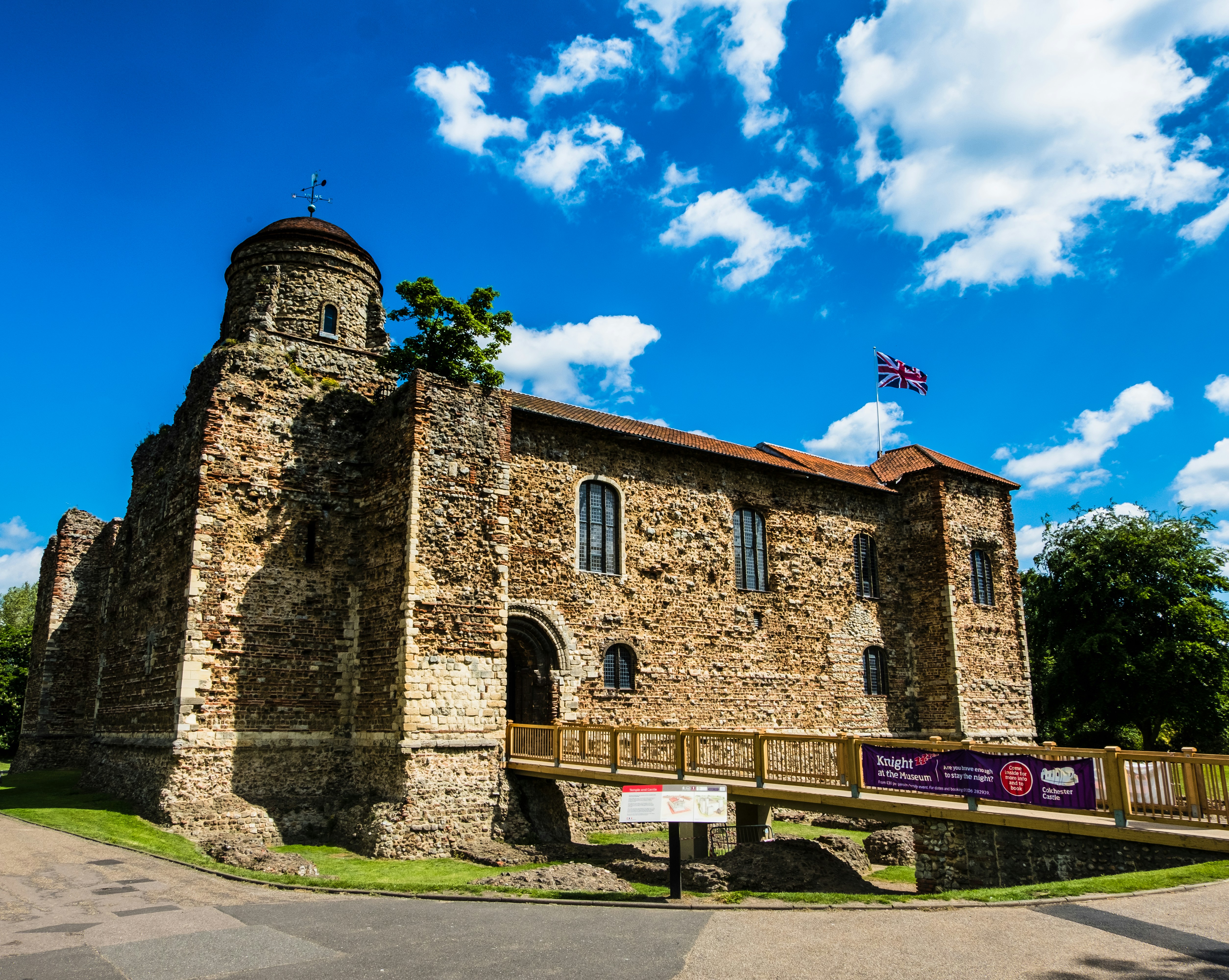 Historic stone fortress with arched doorway and a raised wooden walkway, Union Jack flying atop, under a vivid blue sky with scattered clouds.