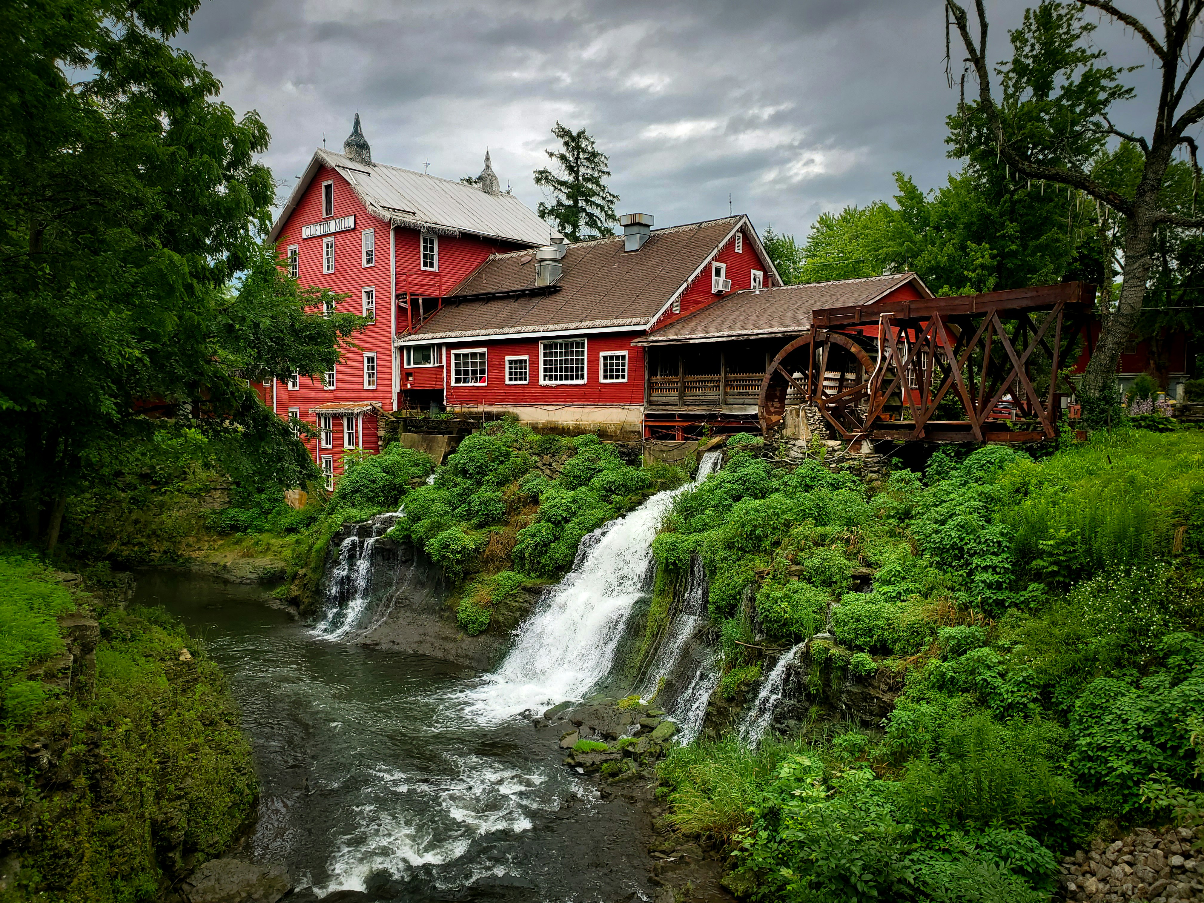 Historic red mill perched above cascading falls amid lush greenery and a wooden wheel bridge.