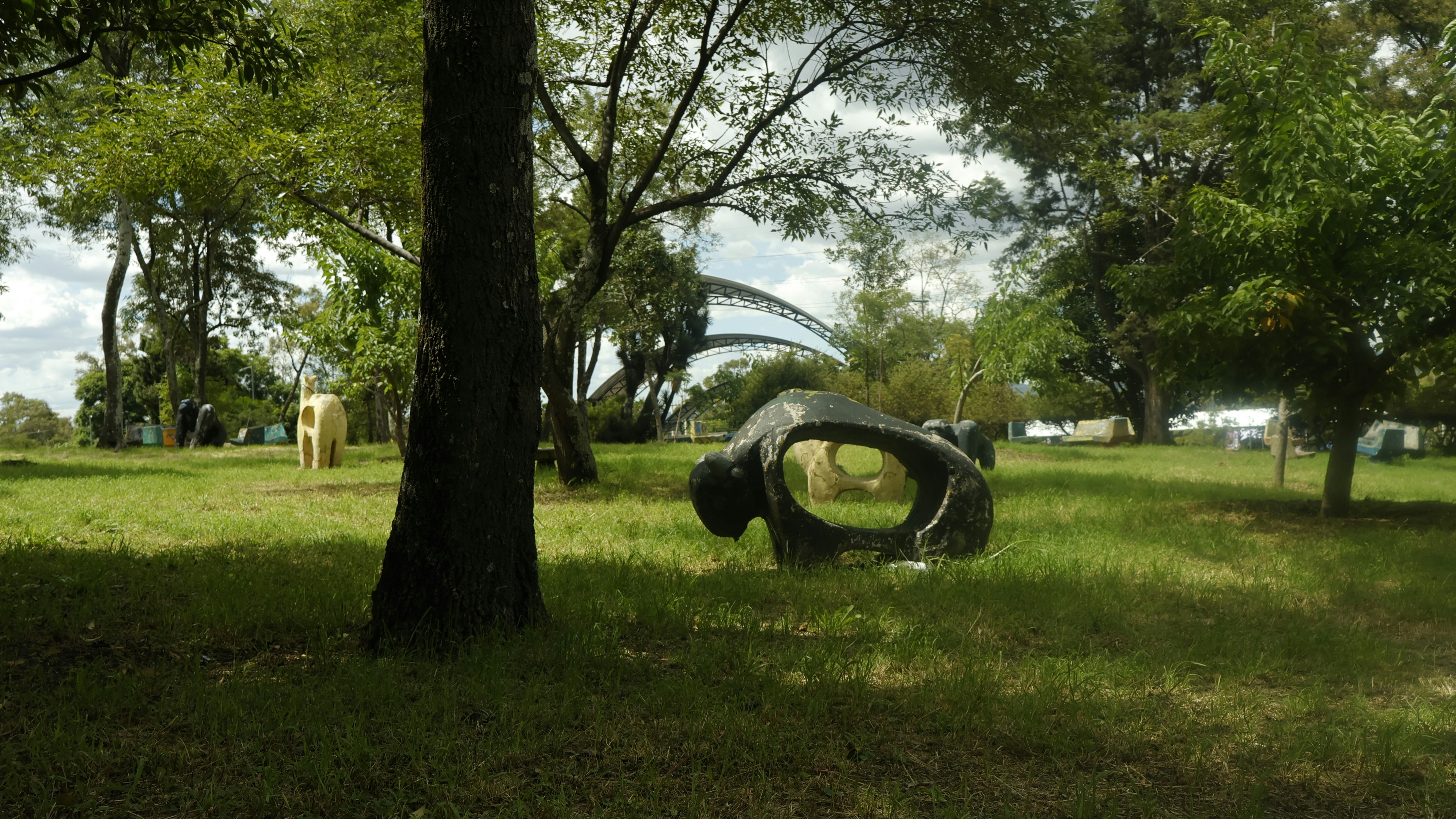 Sculptural form resting on grass among trees in a park with a distant bridge; emphasizes outdoor sculpture and natural light. Note: a small art installation in an open green space.