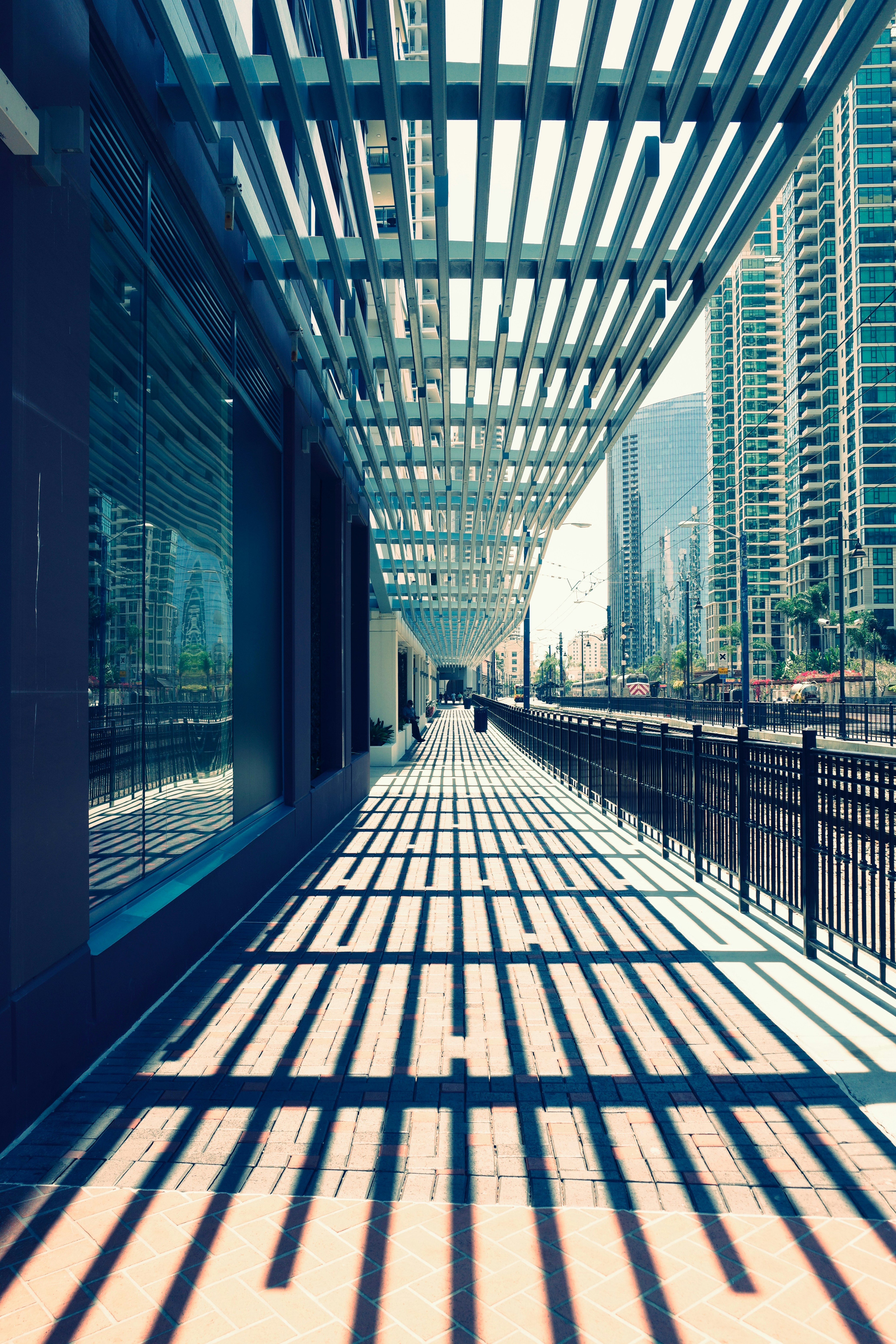 Sunlit walkway beside a glass building, with a lattice overhead casting striped shadows across the brick pavement.