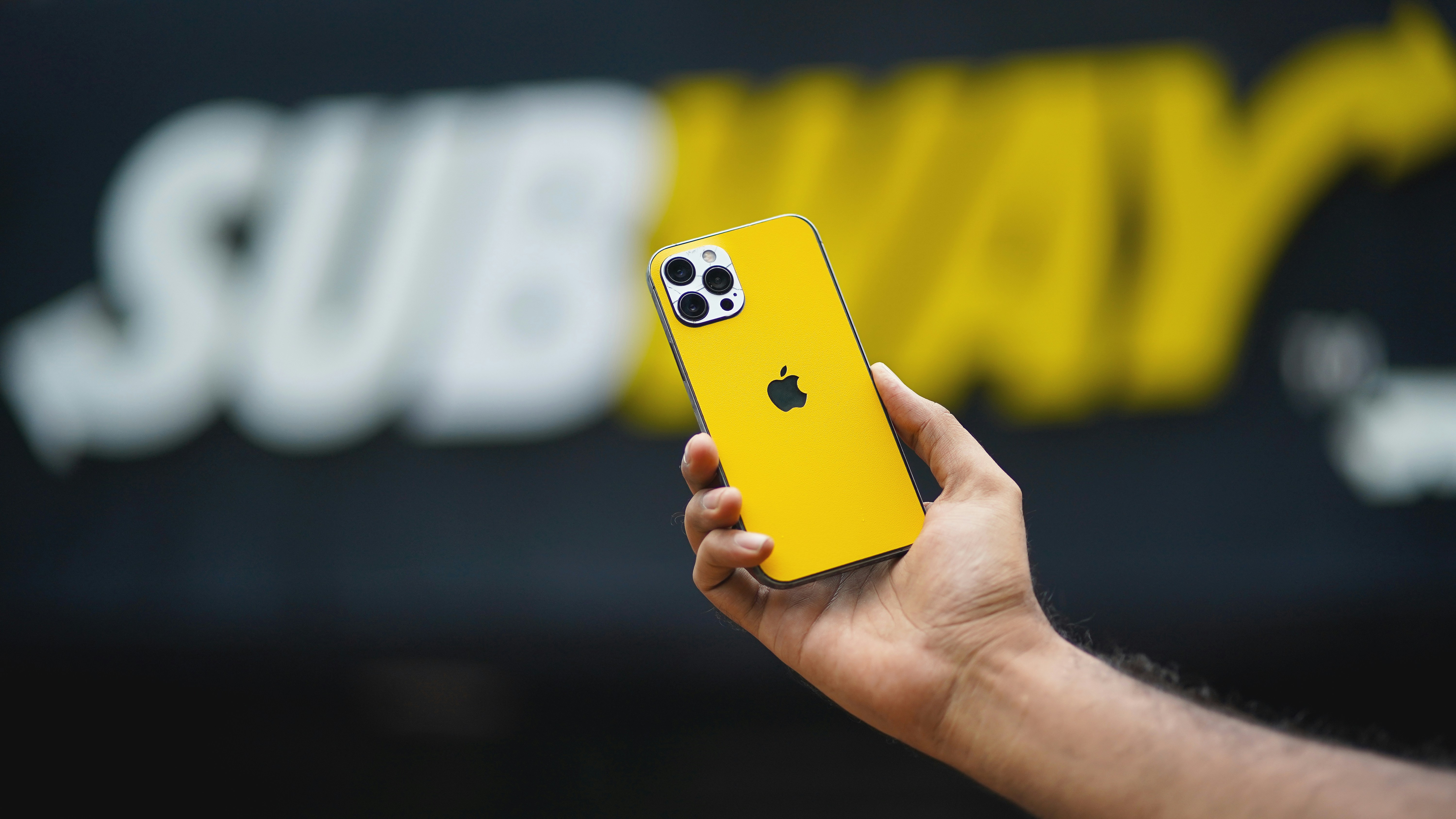 A yellow iPhone held in one hand against a dark, softly blurred background, creating a bold contrast between the vibrant device and the moody surroundings. The shallow depth of field isolates the phone while the generous negative space gives the image a clean, modern commercial aesthetic.