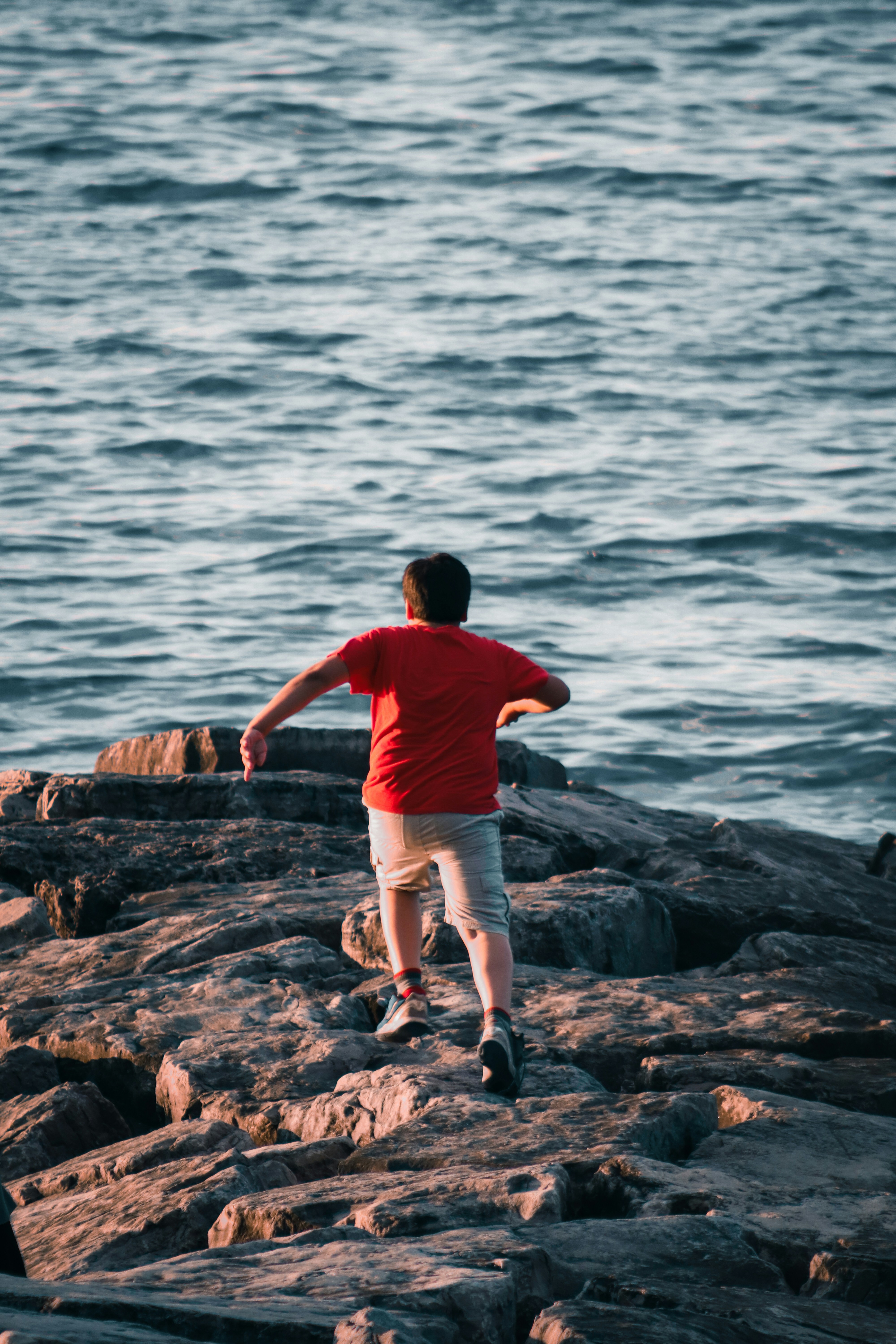 Rearward Ascent: A child in red scales the rocky breakwater toward the open sea
