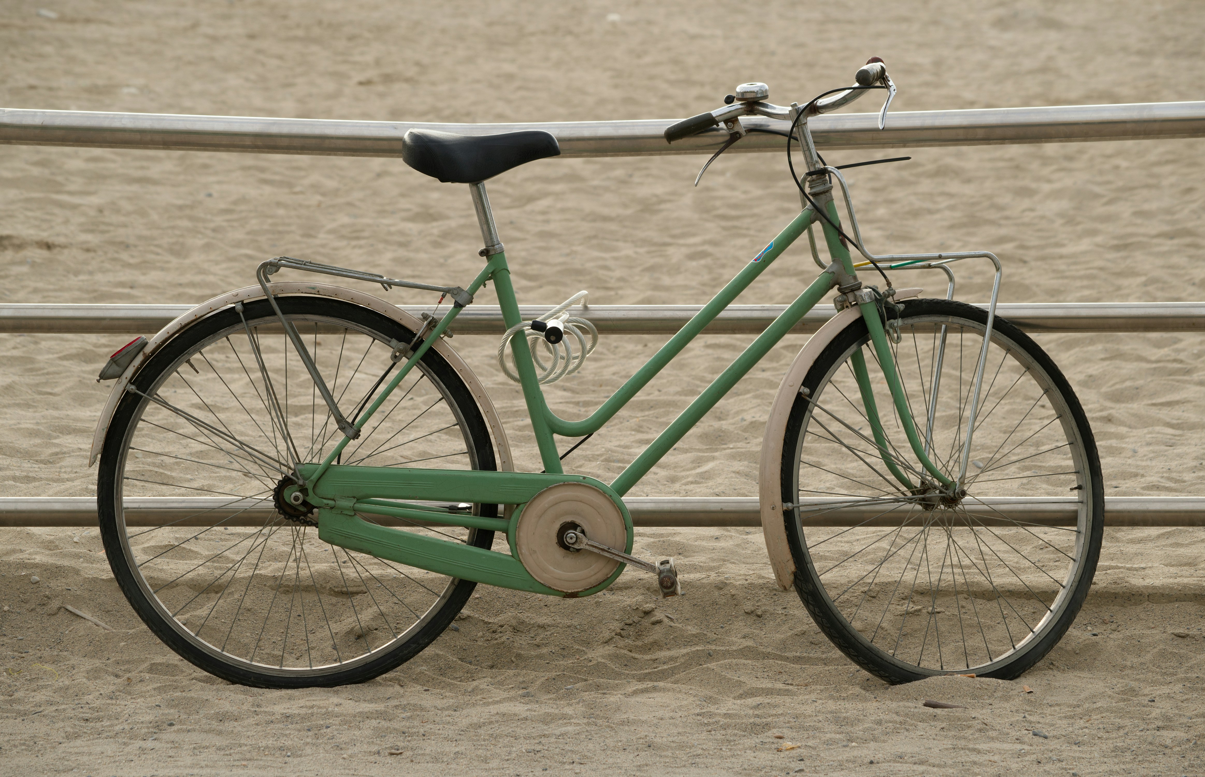 Green vintage bicycle rests beside a railing on a sandy promenade, sunlight softening its paint and details.