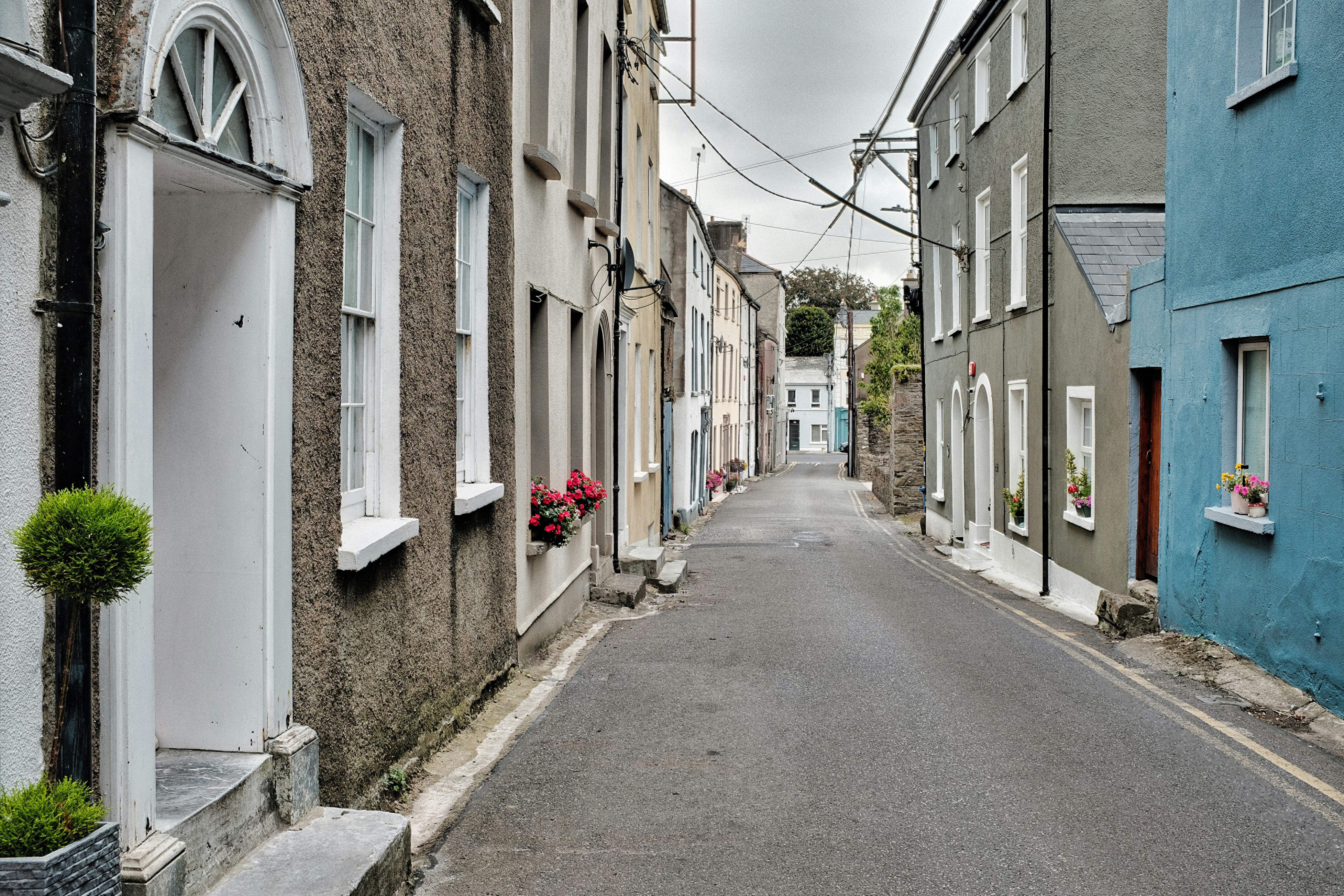 A narrow residential street lined with pastel-hued houses and flower boxes along a grey pavement, receding toward a vanishing point.