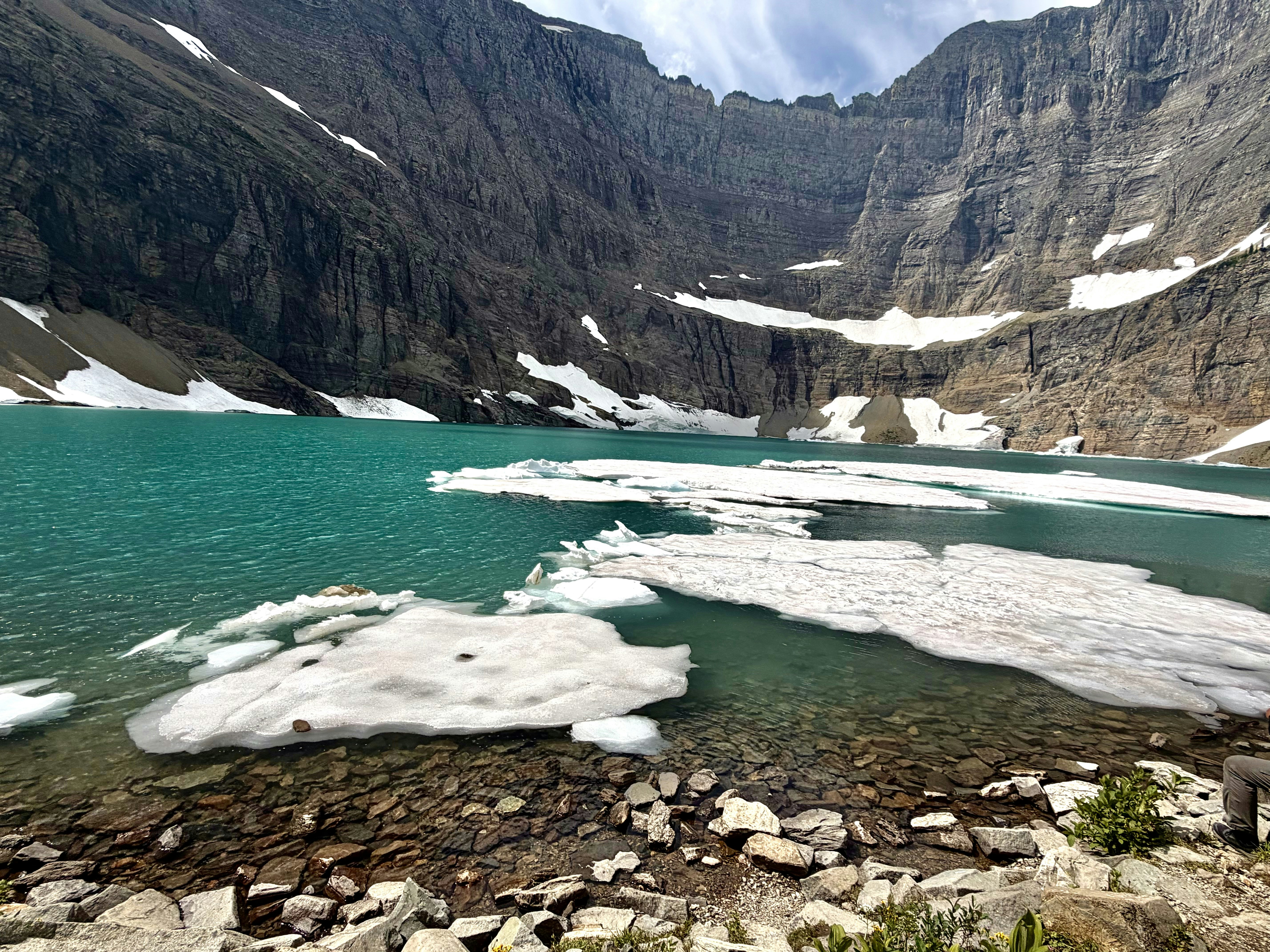 Turquoise alpine lake framed by rugged snow-dusted cliffs and floating ice fragments; rocky shoreline foreground with clear water revealing stones beneath and distant mountain walls.