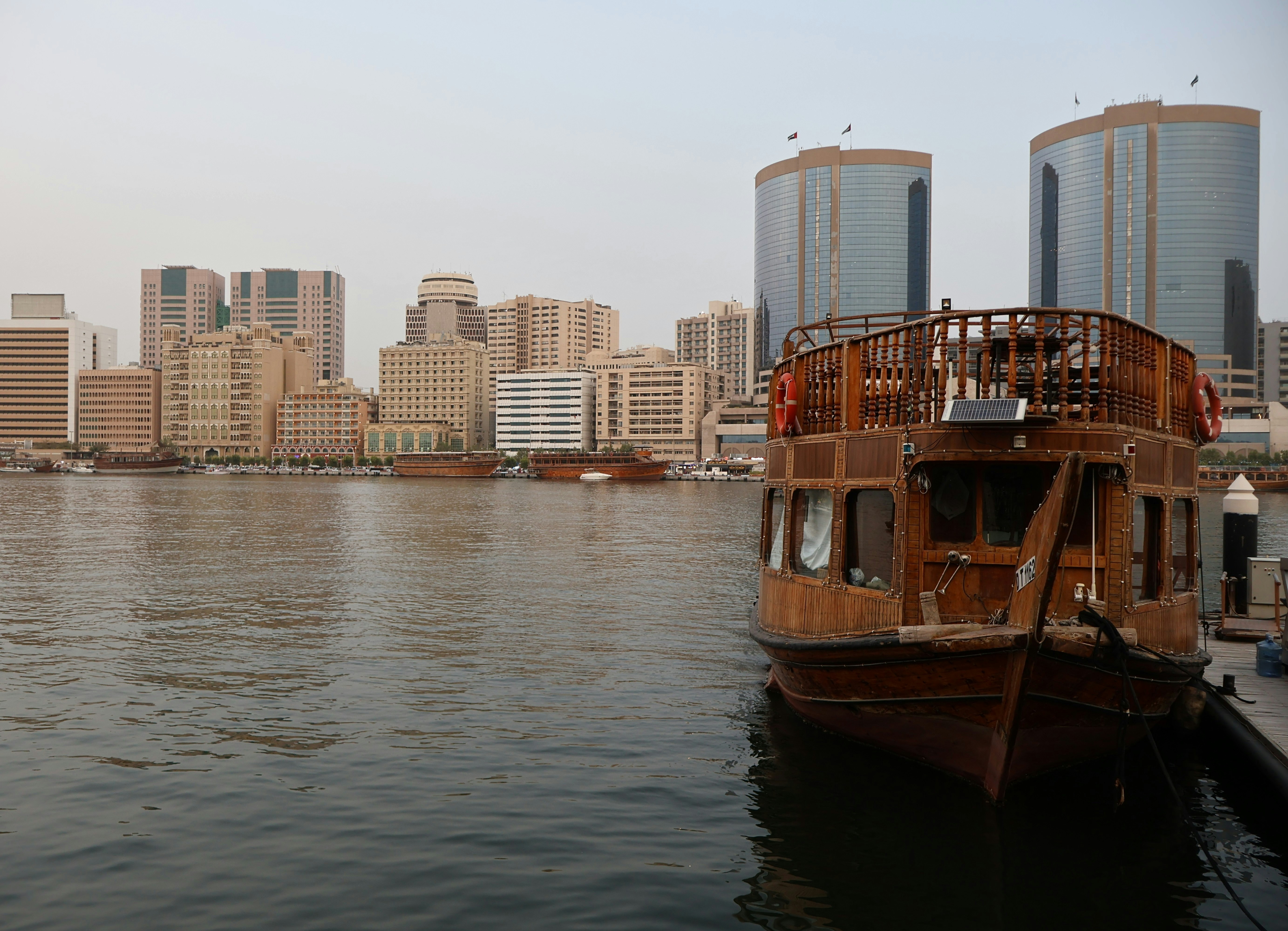 Wooden boat moored at a calm harbor with a city skyline and high-rise buildings in the background.