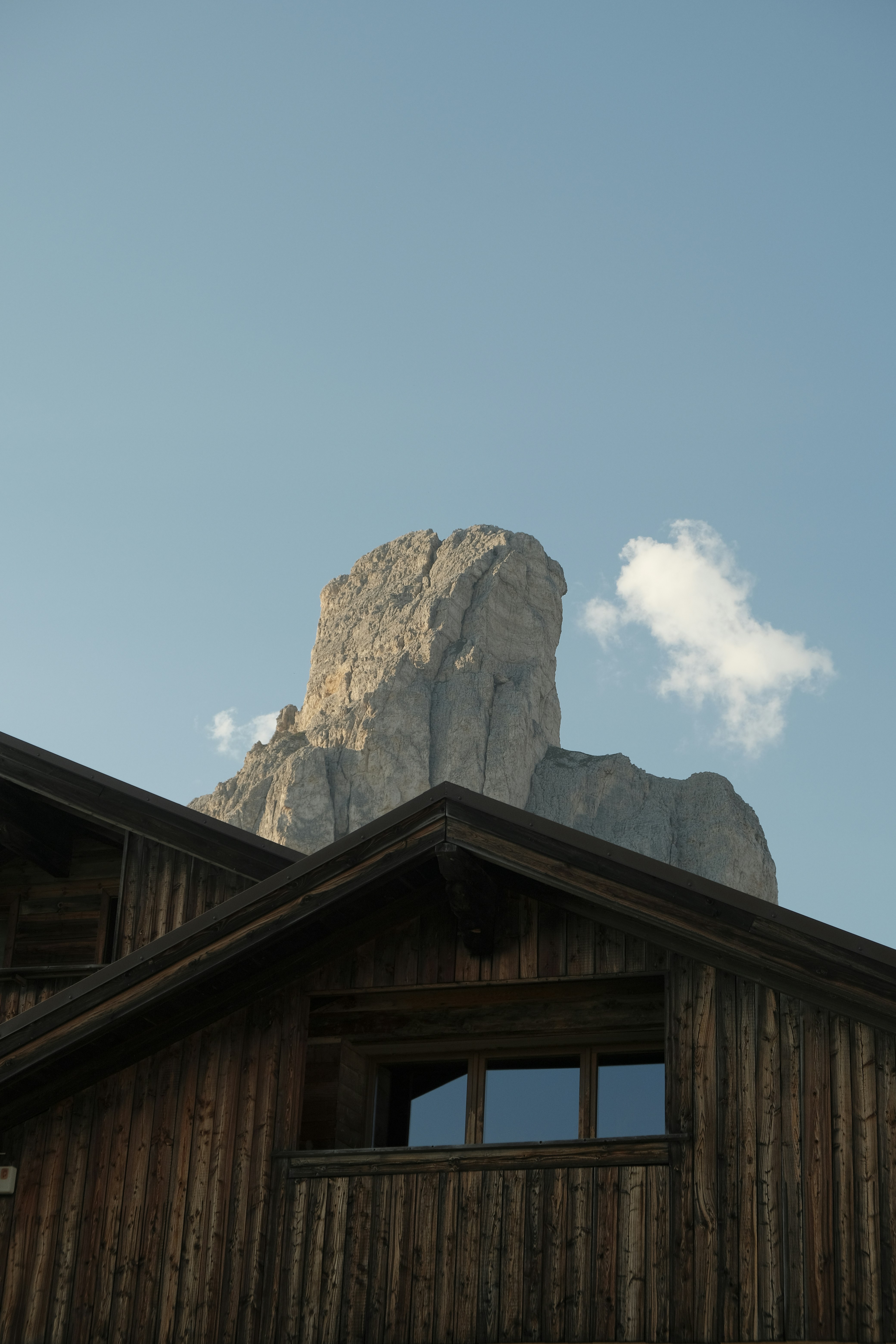 A wooden cabin foreground with a dramatic rocky peak rising behind under a clear blue sky. The scene emphasizes rustic architecture framing a granite summit.