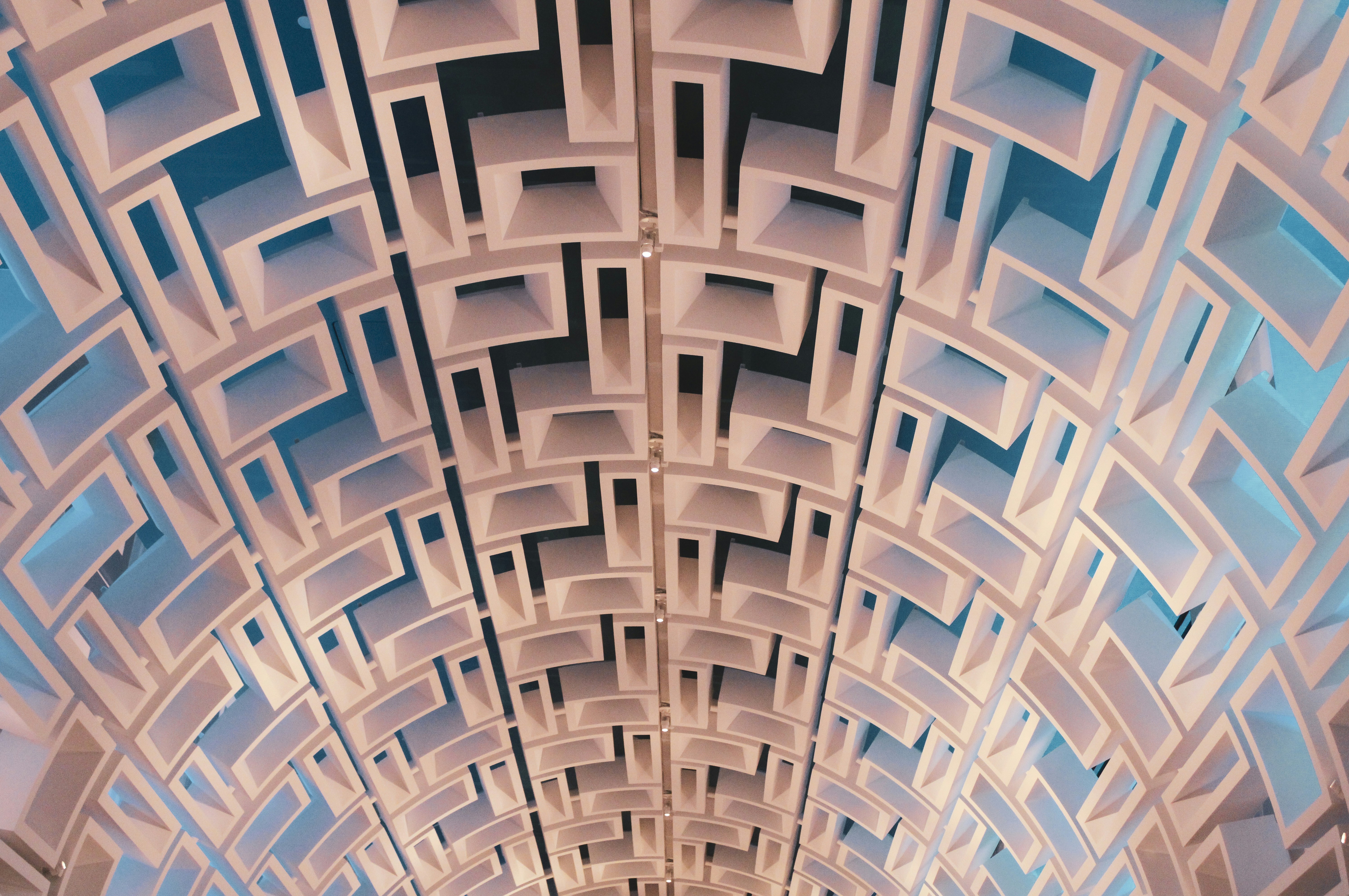 Geometric lattice of rectangular recesses forms a hypnotic ceiling pattern with soft blue and peach tones. The composition emphasizes repetitive depth and architectural texture. This is a photographic capture of an interior ceiling installation.