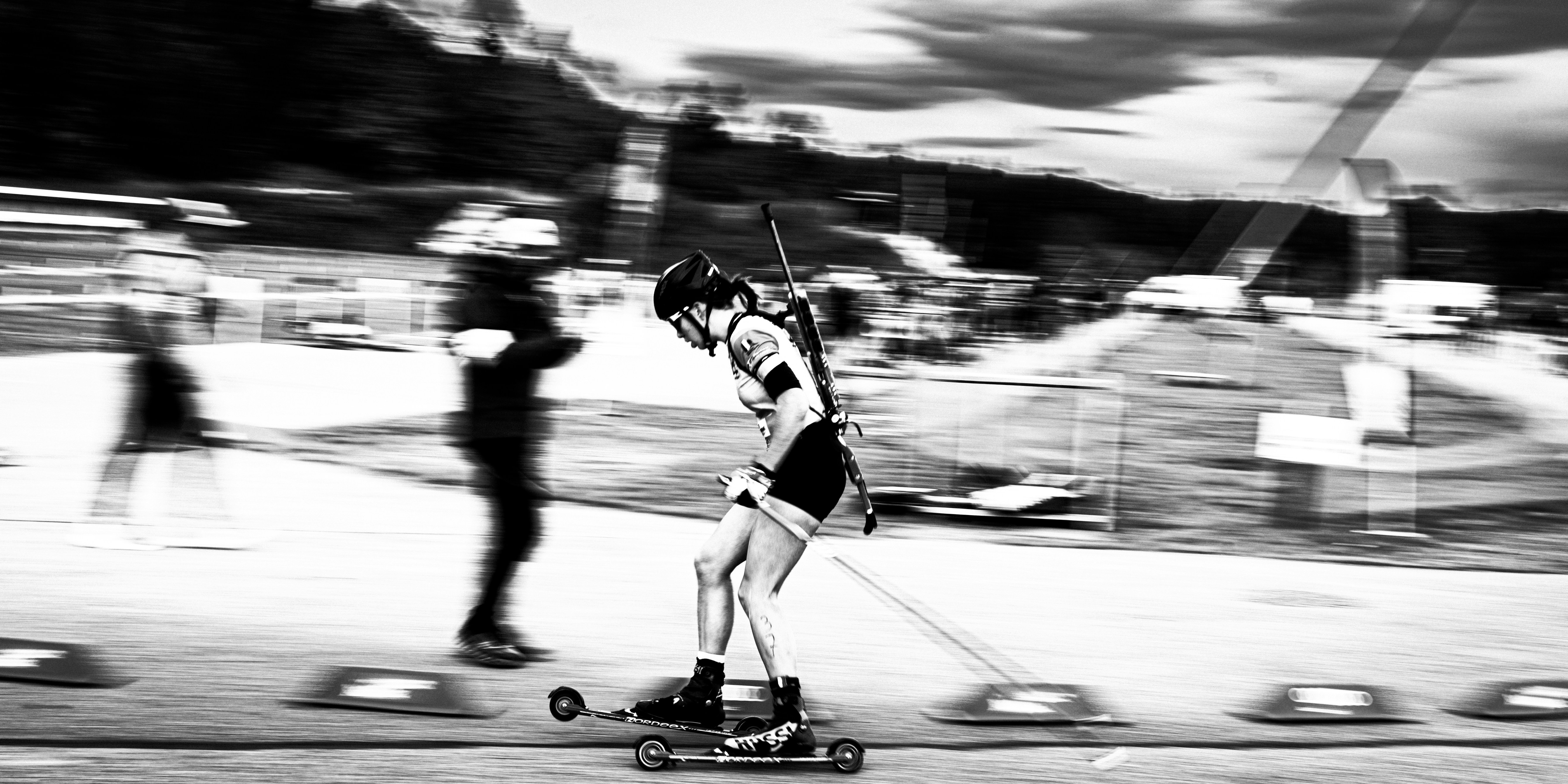 Motion-blurred skateboarder with inline gear along a racecourse; crowd and banners form a dynamic backdrop, emphasizing speed and sport in an urban setting.