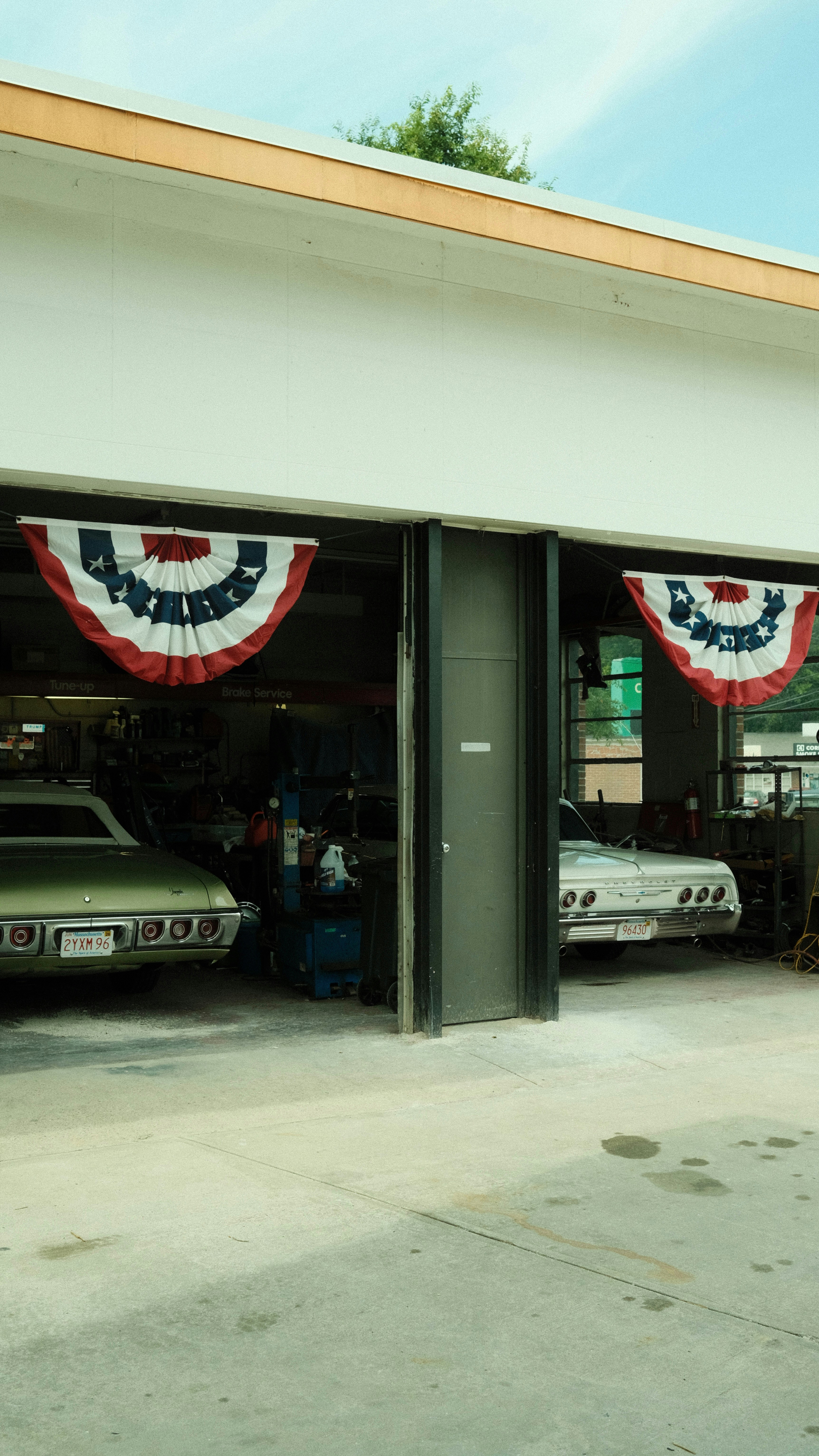A classic car service garage with two open bays, red-white-blue bunting, and vintage cars inside. The scene conveys a retro automotive vibe and workshop activity.