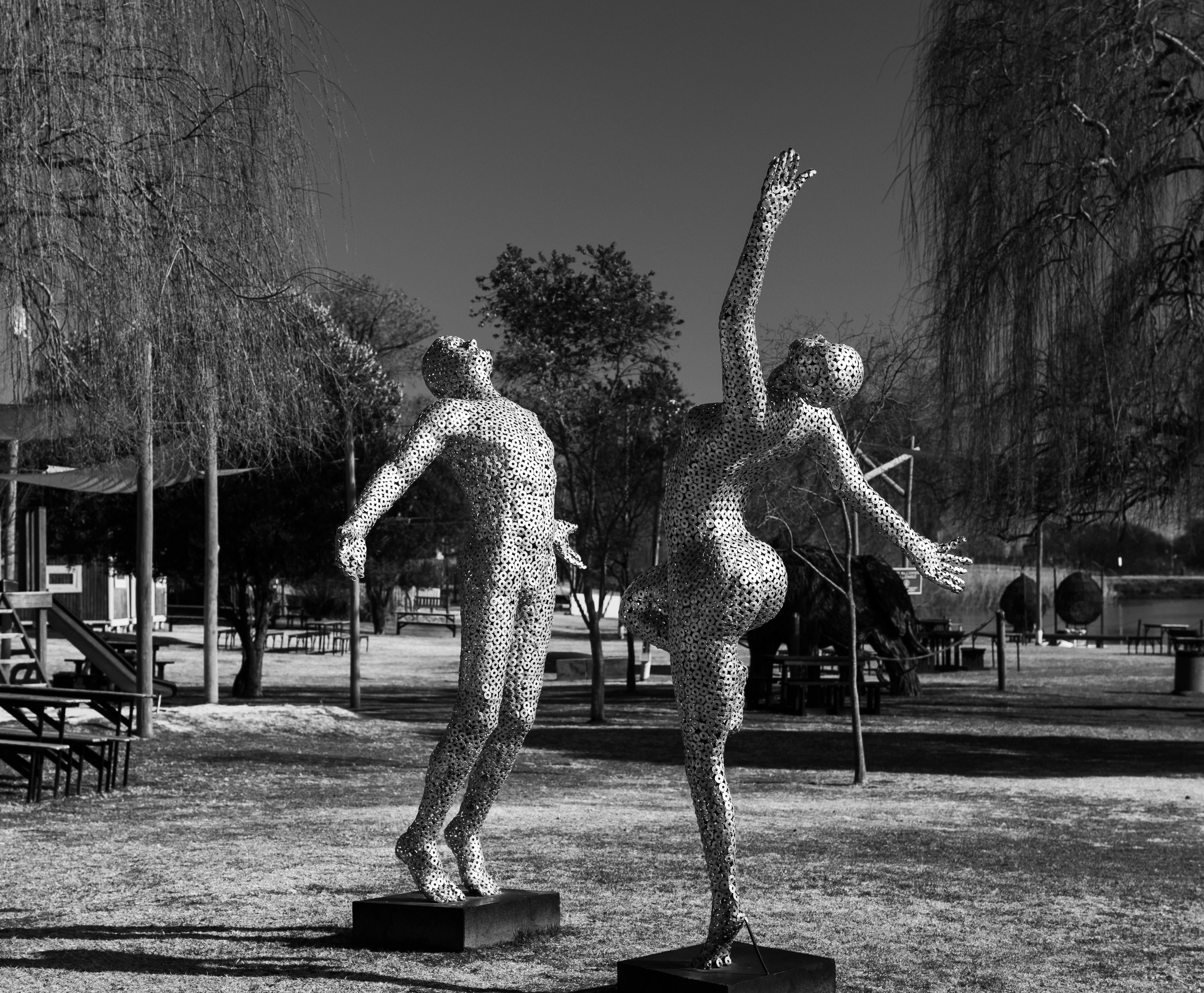 Two metallic statues dance in a sunlit park, their reflective surfaces catching afternoon light amid trees and benches.