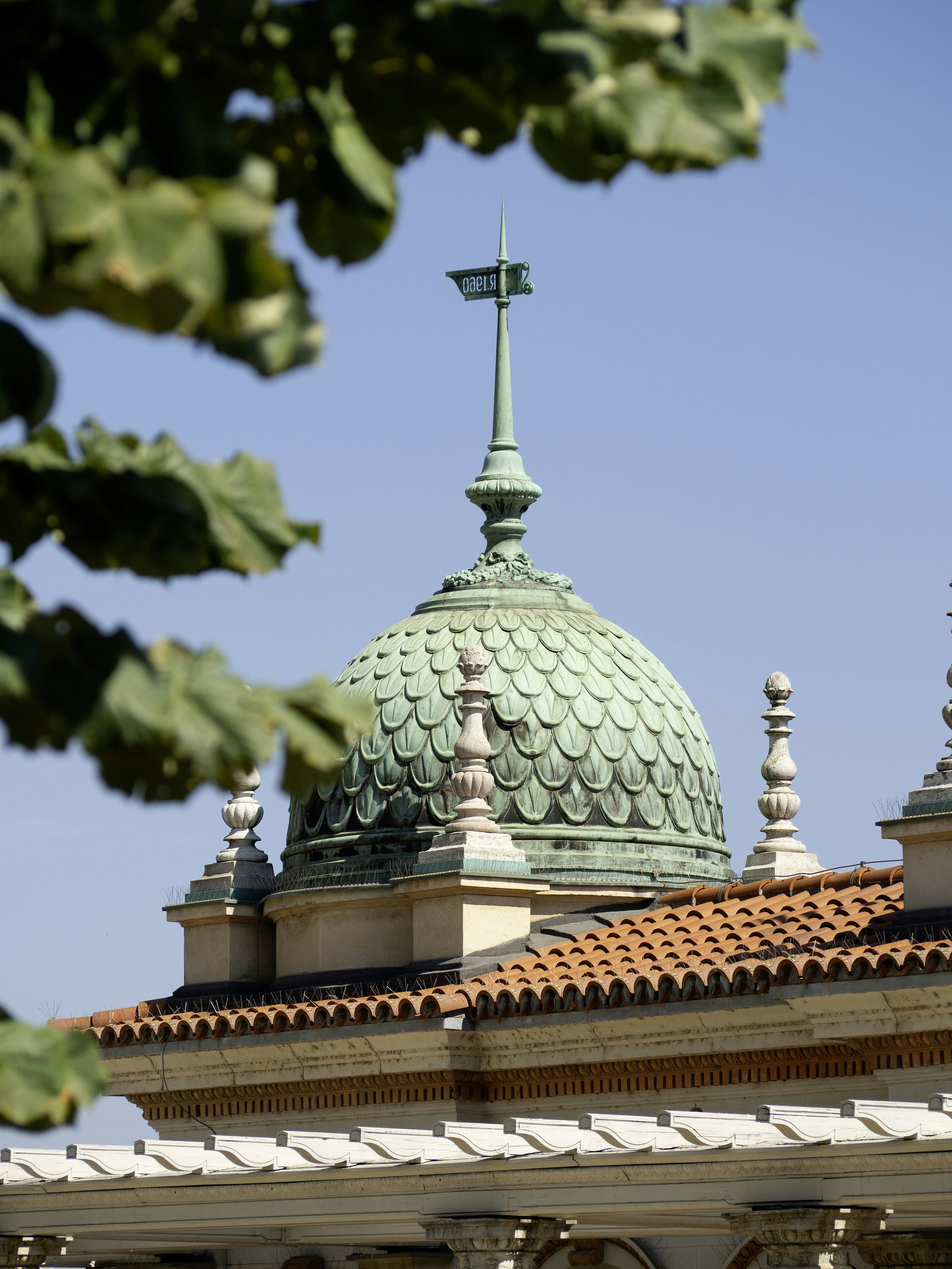 Architectural dome with green patina and red-tiled roof accent under a blue sky, framed by leaves in the foreground. The shot emphasizes the dome's ornate details and rooftop ornaments.