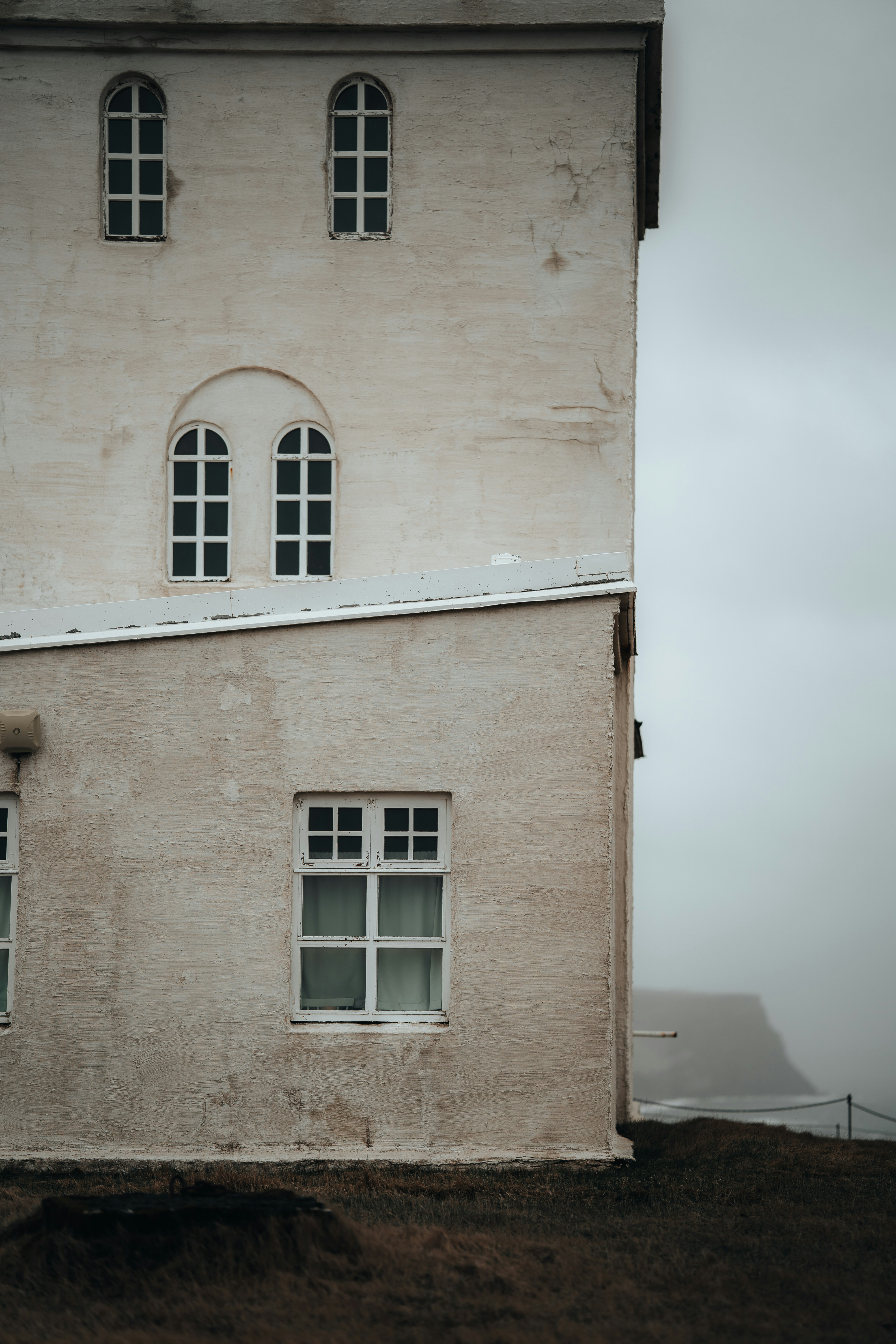 Brick-built structure with arched and rectangular windows, set against a foggy seascape; a solitary building edge dominates the left foreground and the right fades into misty horizon.