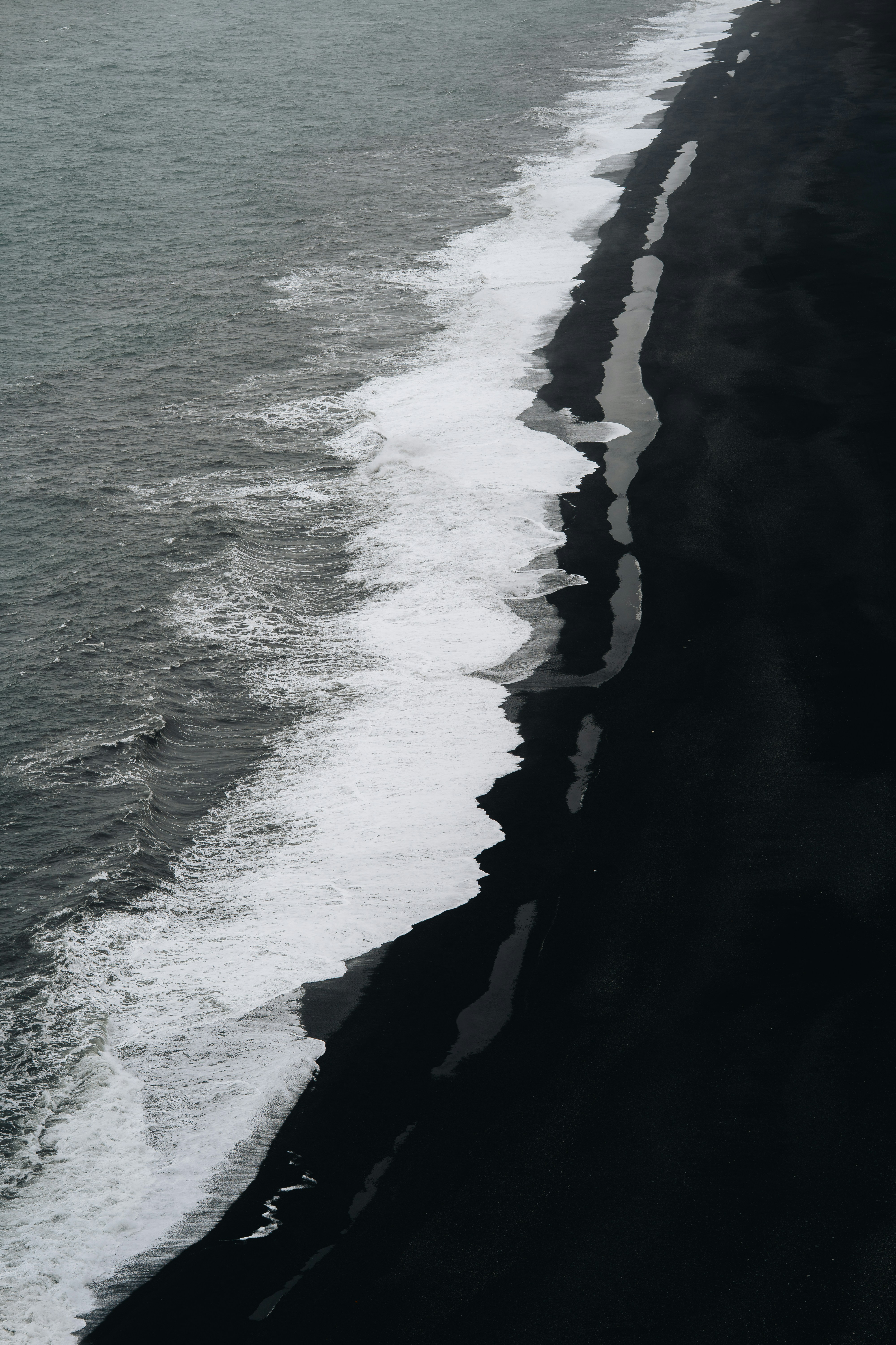 Monochrome coastal scene showing a dark volcanic beach meeting white-capped breakers along a jagged shoreline.