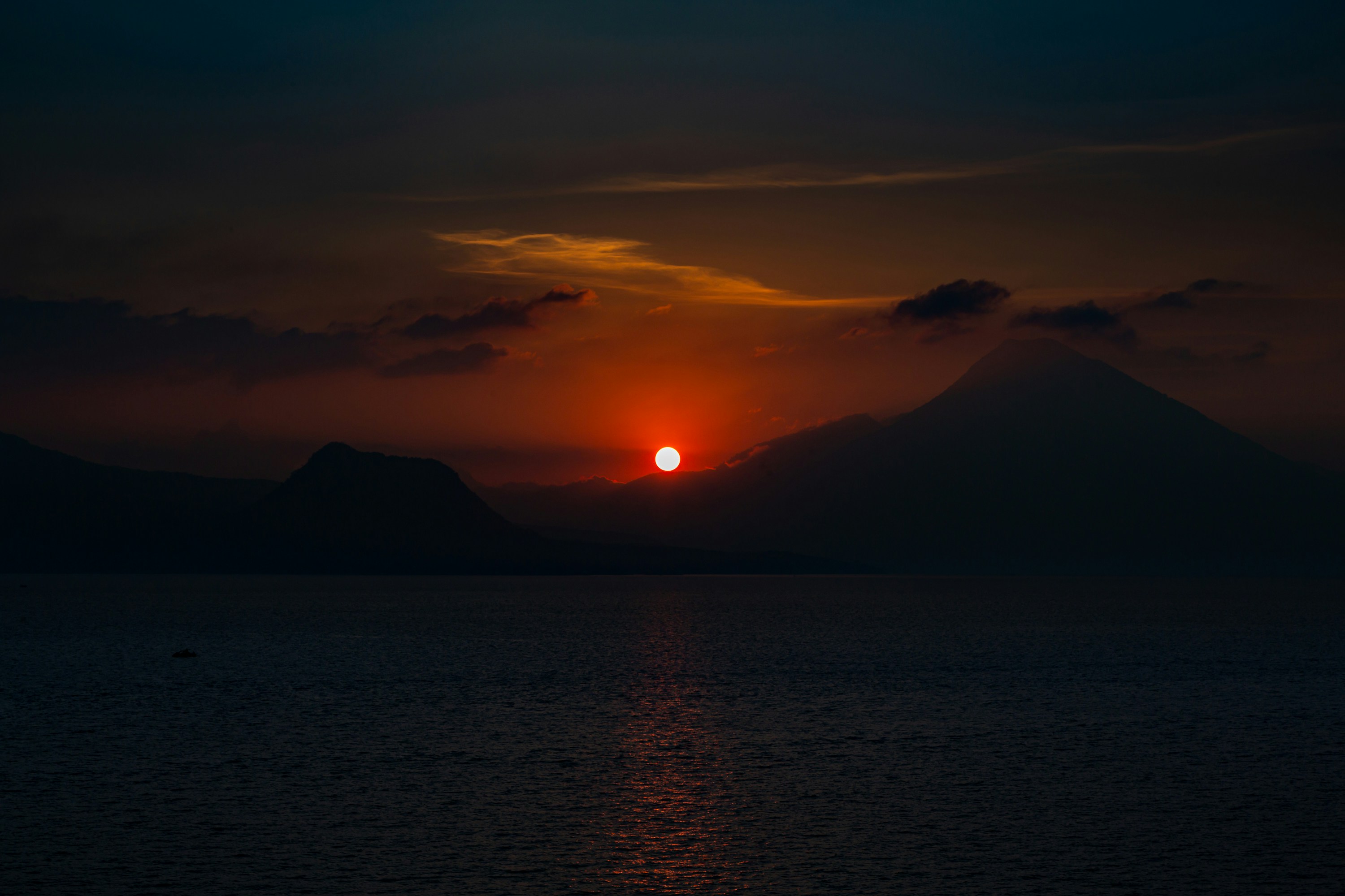 Low, warm sun sinks behind distant silhouettes as calm water mirrors a crimson sky; a serene seascape emphasizing the horizon and glow of sunset.