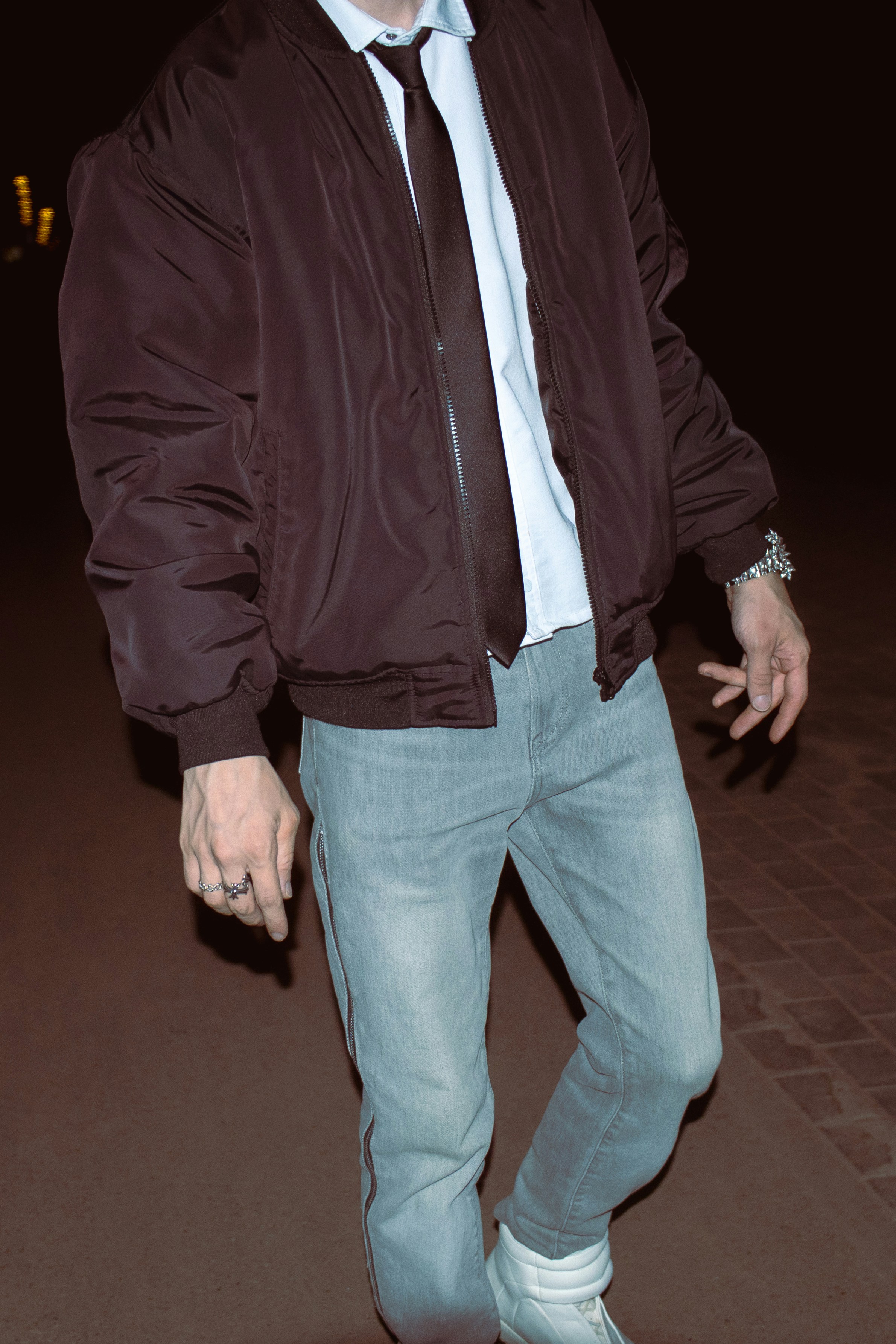 Man in a maroon bomber jacket, light shirt and tie walking on a dimly lit street; hands visible, casual jewelry, jeans and white sneakers. Photograph captures street-night style detail.