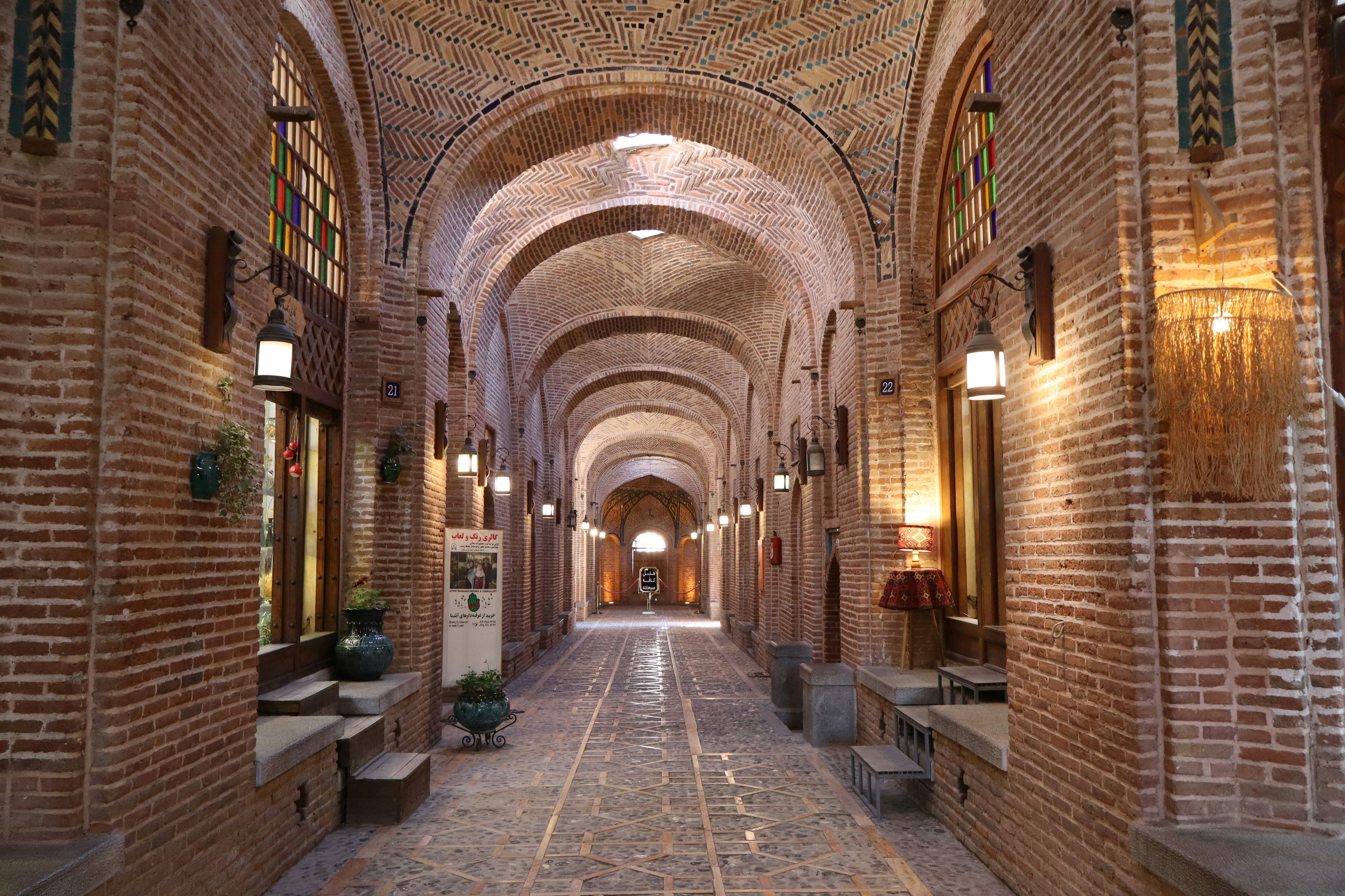 Brick-vaulted corridor with arched ceilings and warm lighting; a long, brick-paved passage flanked by benches and doorways, receding toward a bright doorway at the far end.