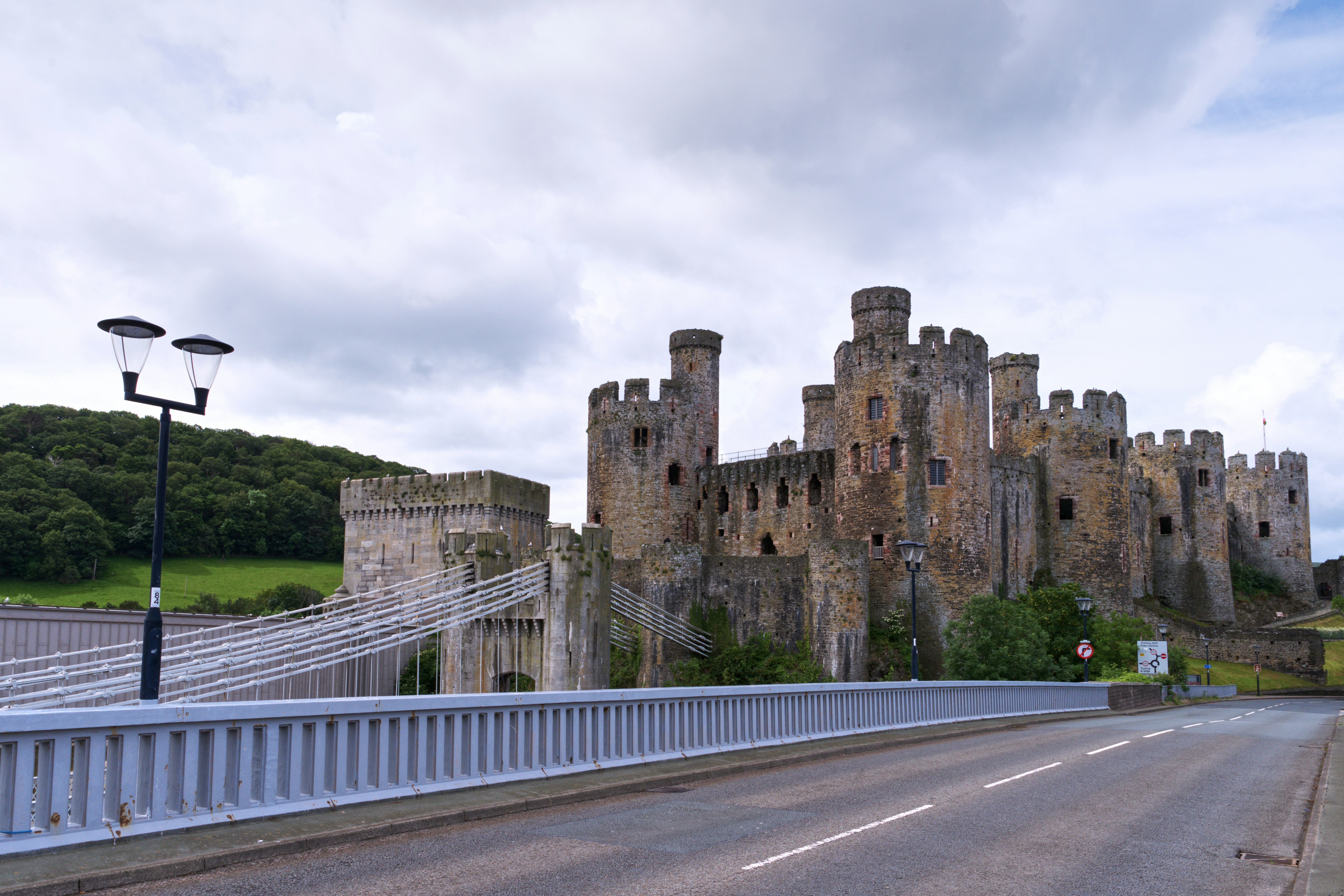 Weathered castle ruins ascend above a quiet river road, with a modern railing and lamp posts guiding the foreground toward the ancient stonework.