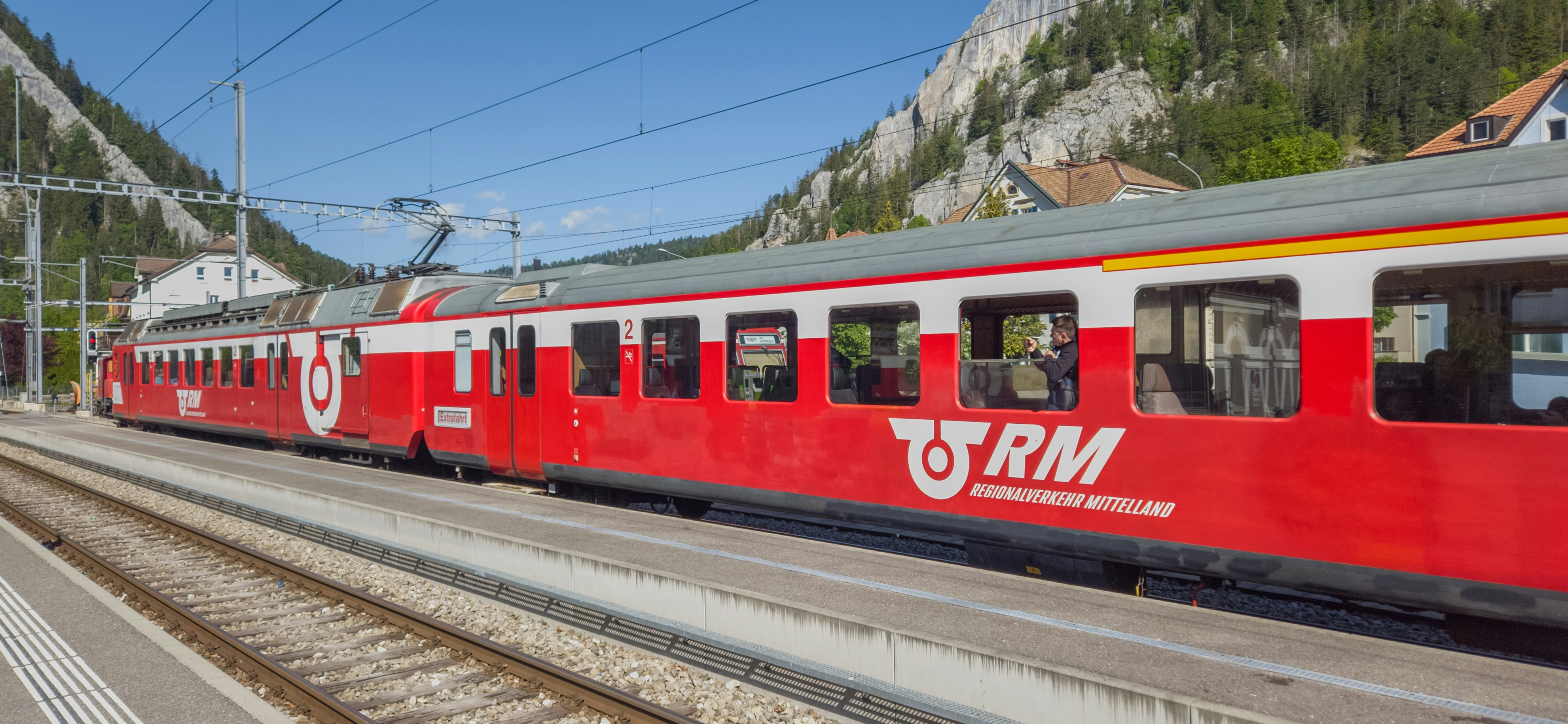 Bright red RM train glides along a sunlit alpine track with mountains in the background and a clear blue sky.