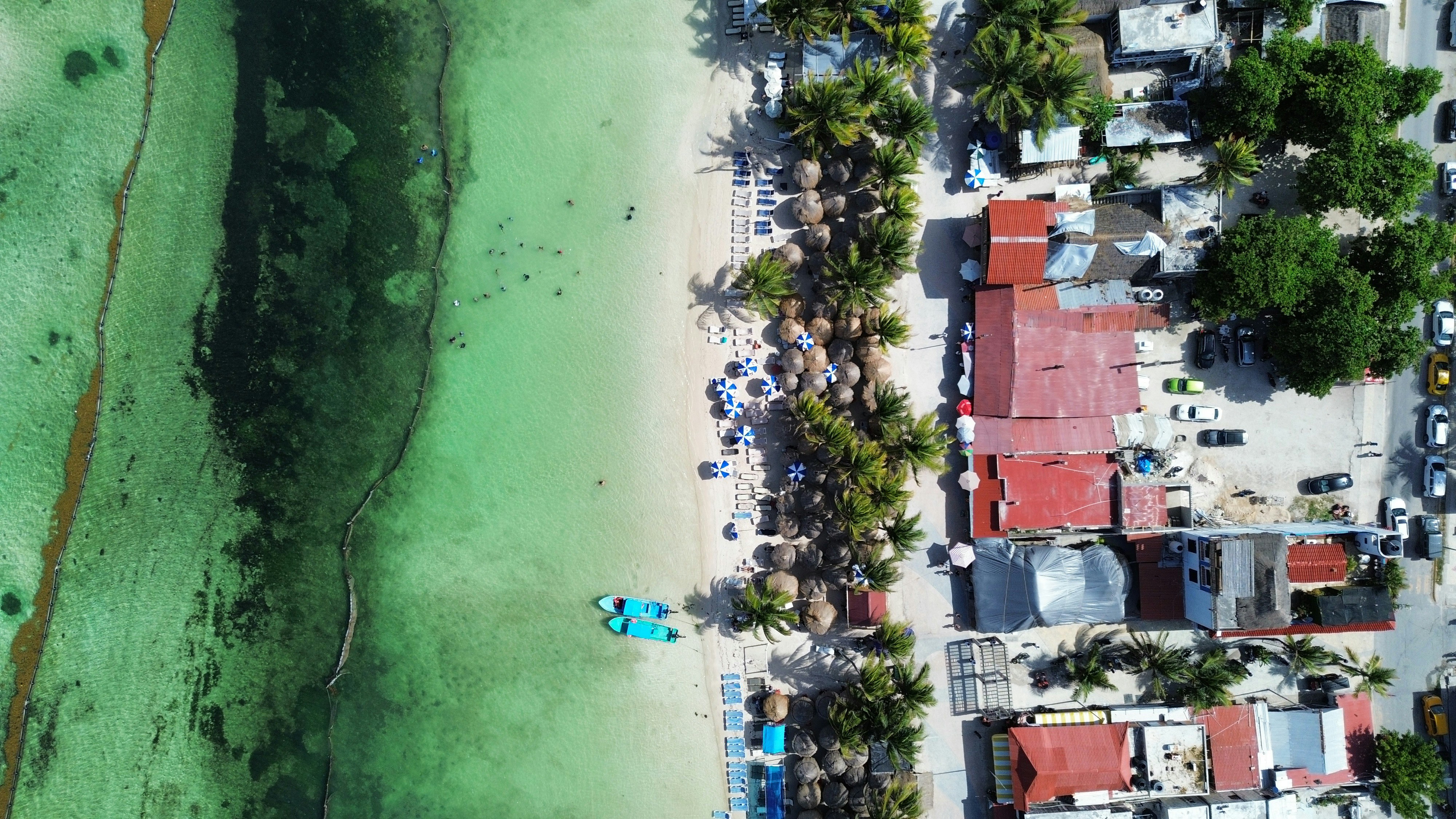 Aerial view of a turquoise-green shoreline with a sandy beach and a row of colorful buildings and market stalls along the coast.