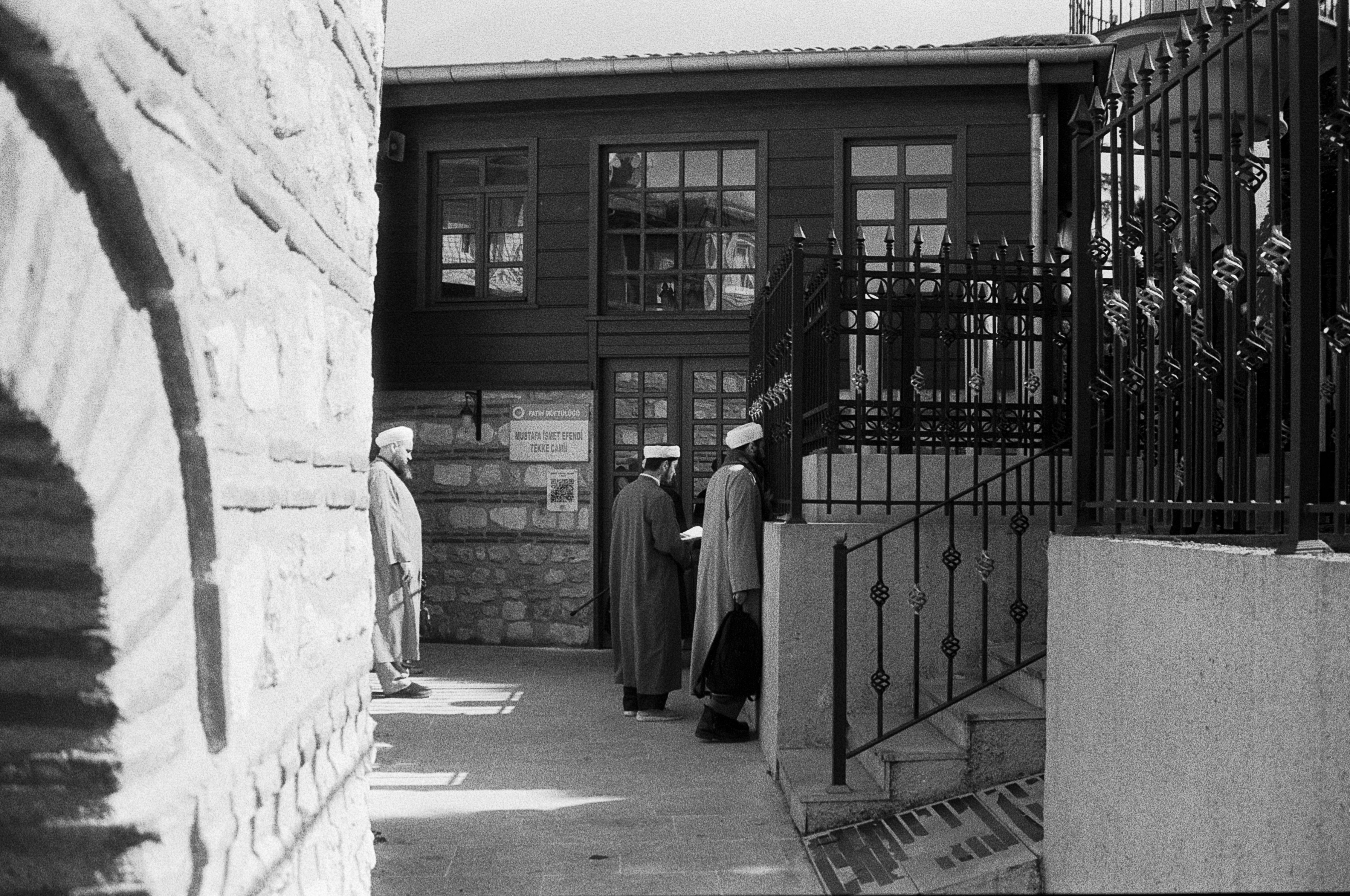 A monochrome scene shows a small group of people in robes gathered near a building courtyard, framed by architectural stonework and a wrought-iron railing; a logo or sign may appear on the wall in the background.