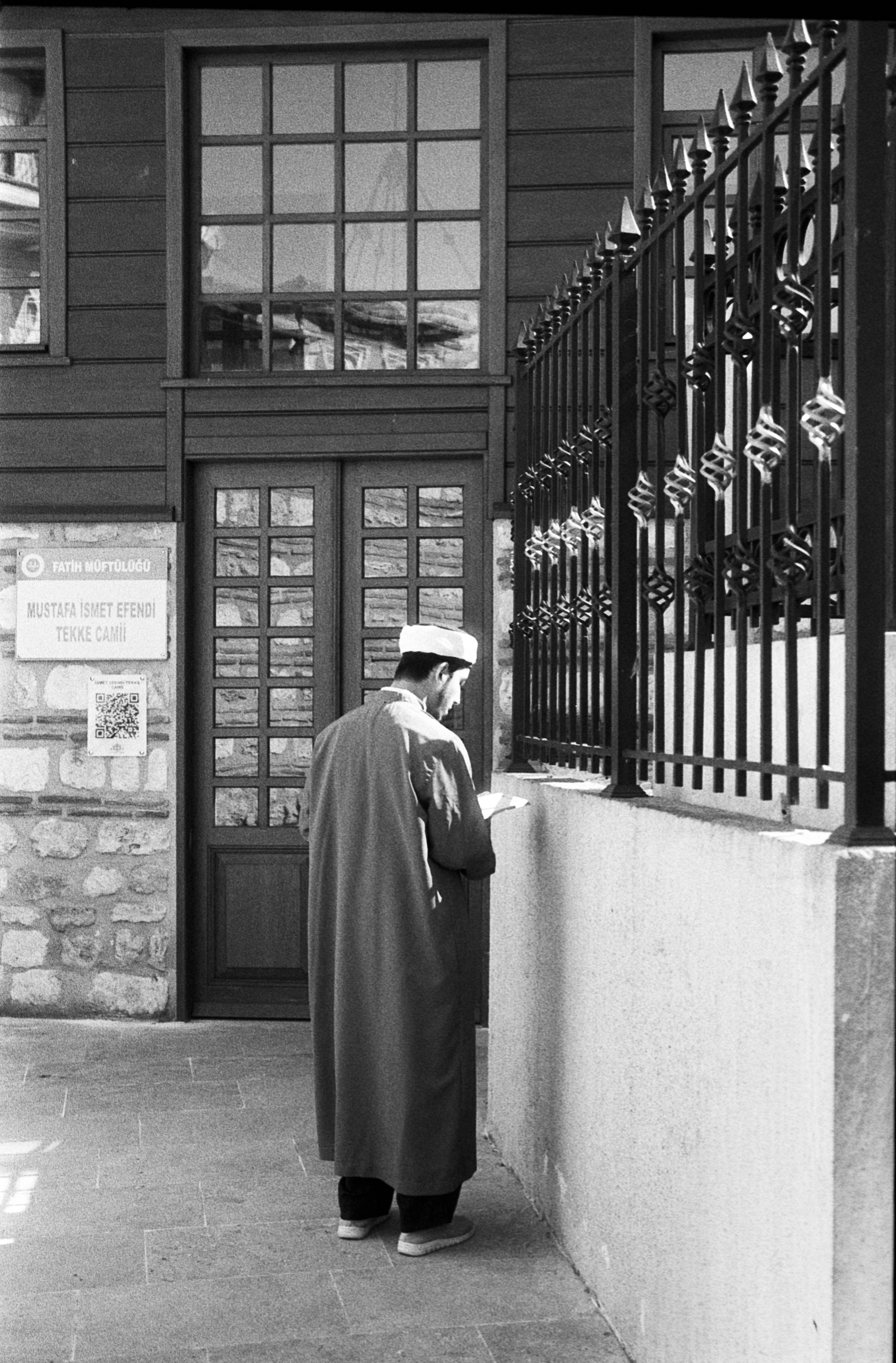 A man in traditional attire reads at a low concrete wall beside a wrought-iron fence and a wooden door, captured in grayscale with strong verticals and subtle textures.