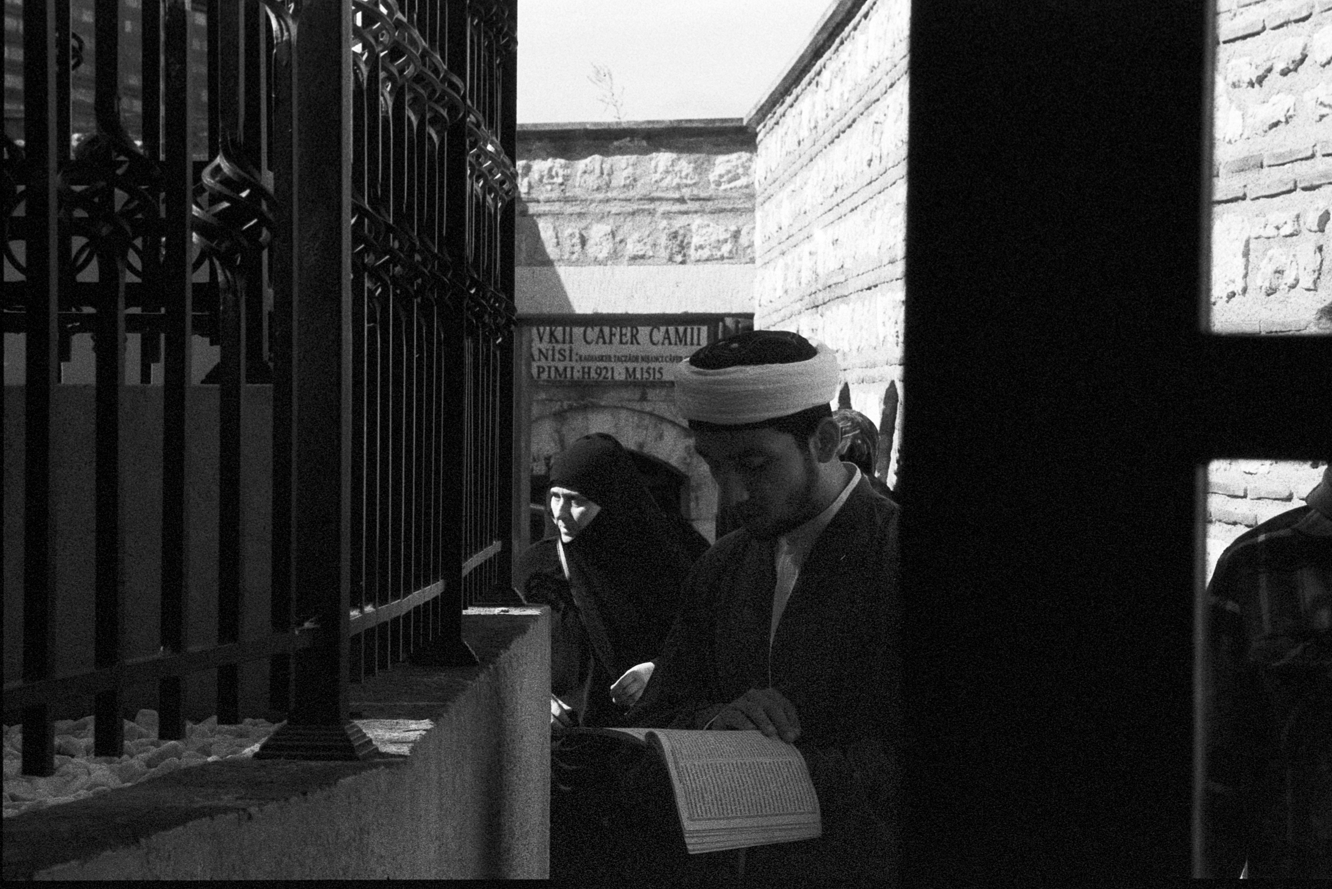 A man wearing a turban reads a newspaper near a gated stone wall, bathed in soft light with another figure in the background; a candid moment with architectural shadows.