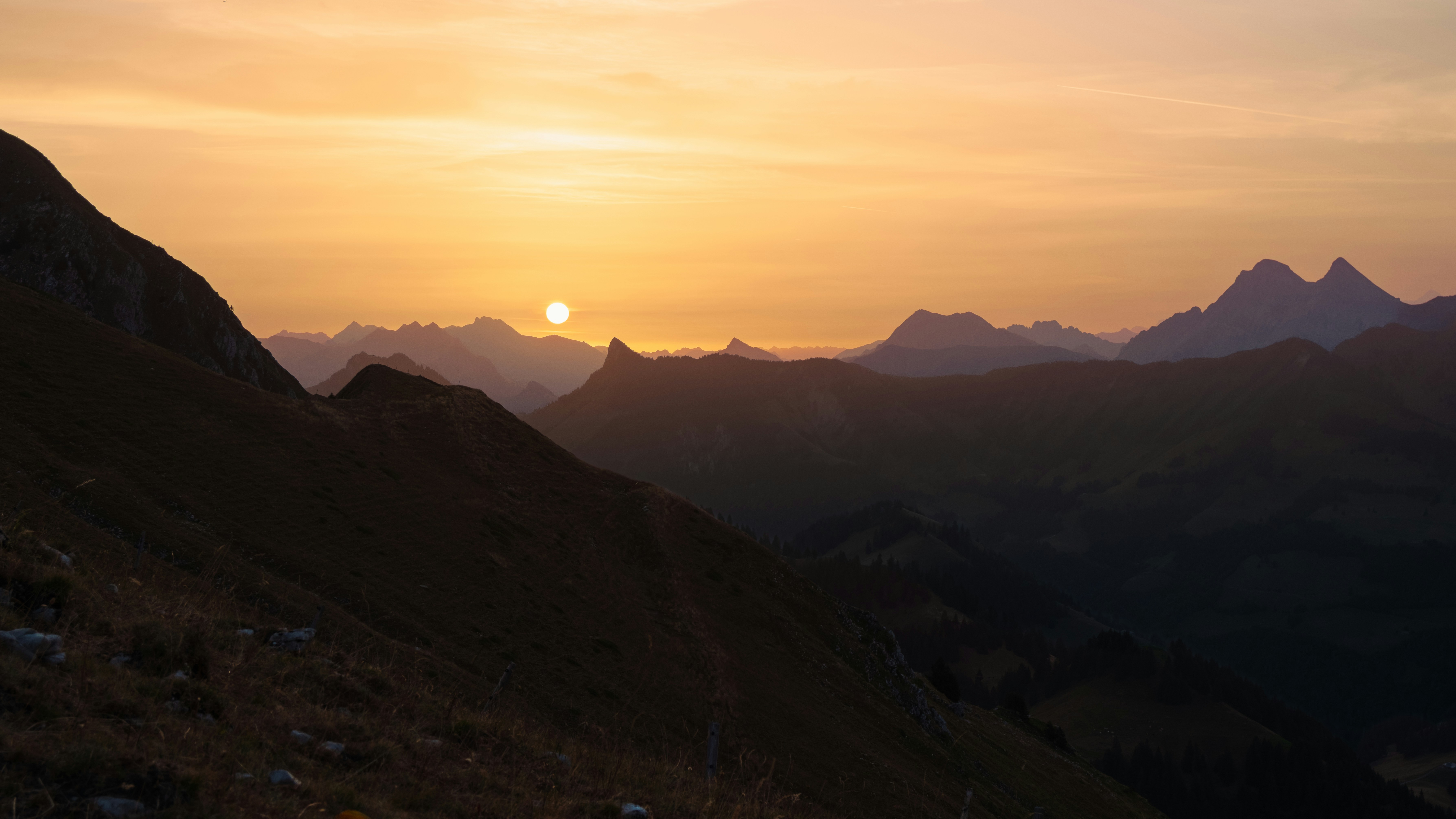 A tranquil alpine landscape at sunset with a soft orange sky and silhouetted mountain ridges.