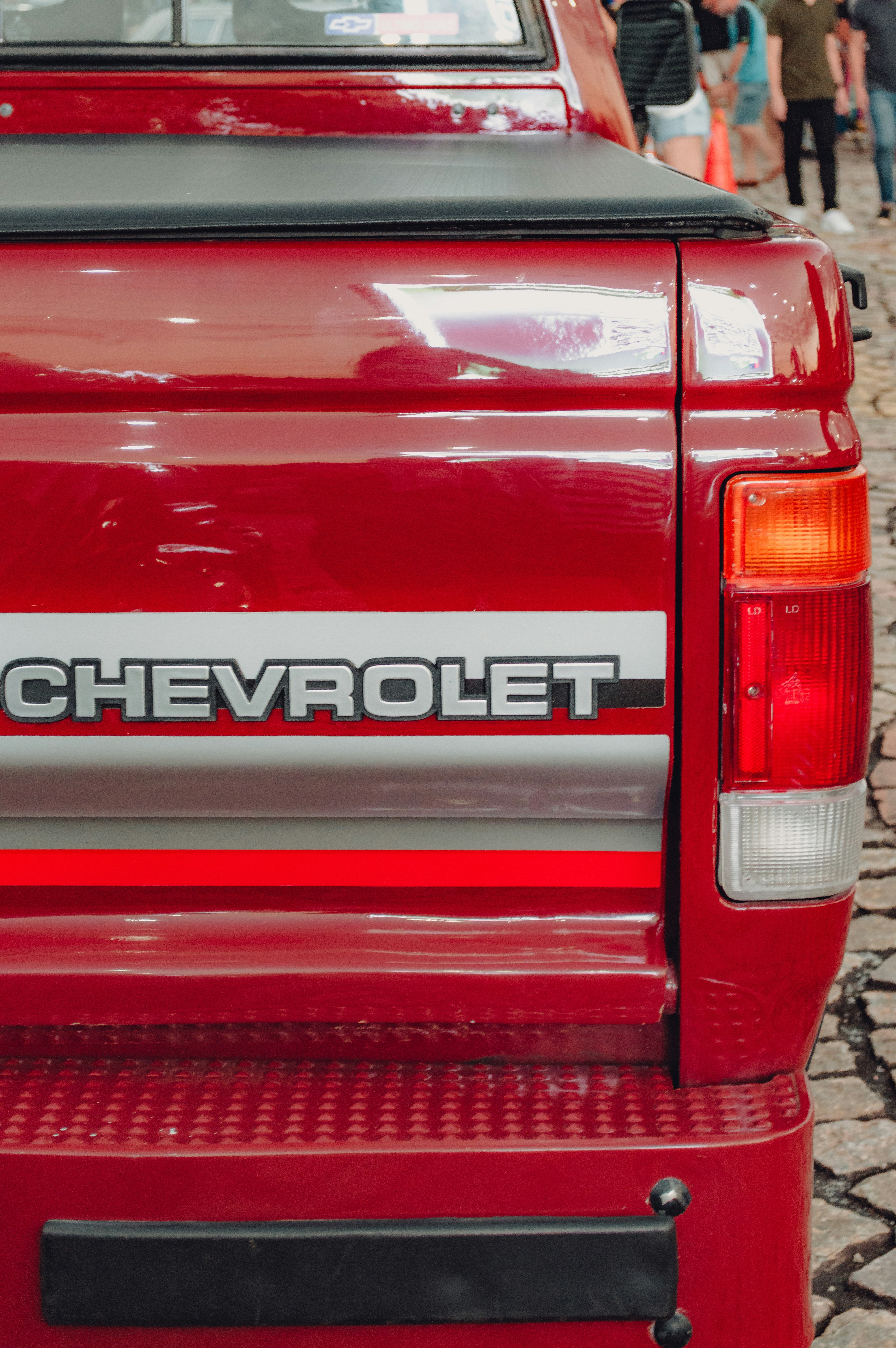 Rear view of a red Chevrolet pickup showcasing the chrome badge against a black bumper and red body with a bright taillight. The scene includes cobbled ground and blurred people in the background, indicating an outdoor event or gathering.