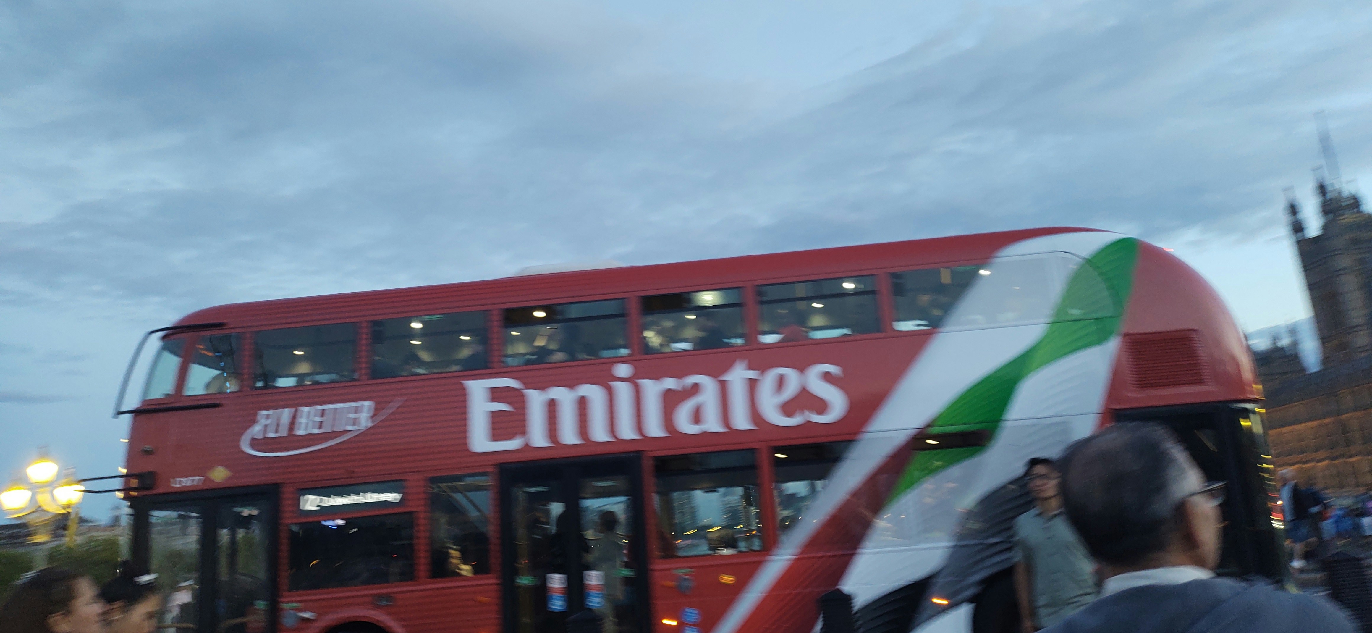 Bright red double-decker bus branded Emirates with pedestrians nearby in a busy urban setting.