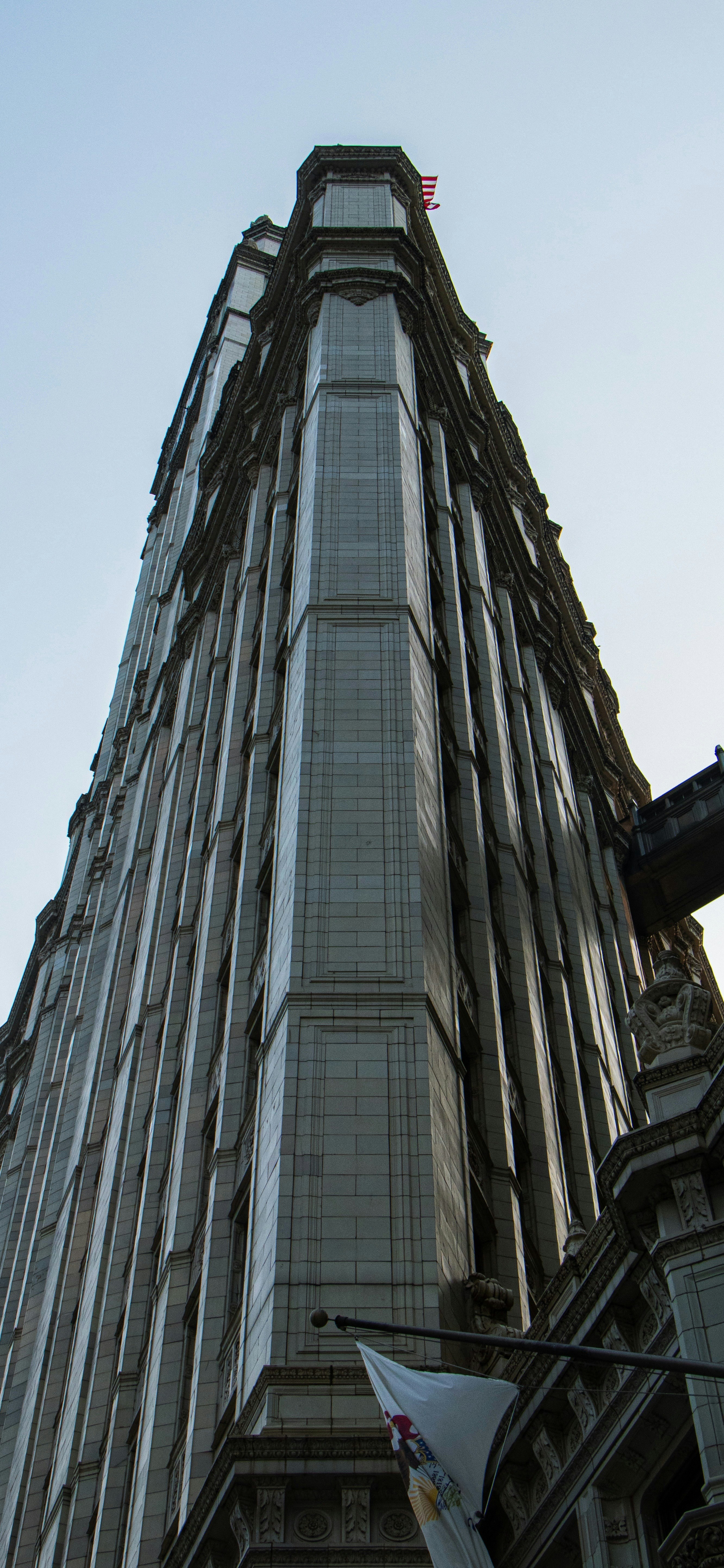 Tall, ornate building towers upward with glass and stone detailing against a clear sky; foreground shows a flag and architectural cornices indicating a grand urban facade.